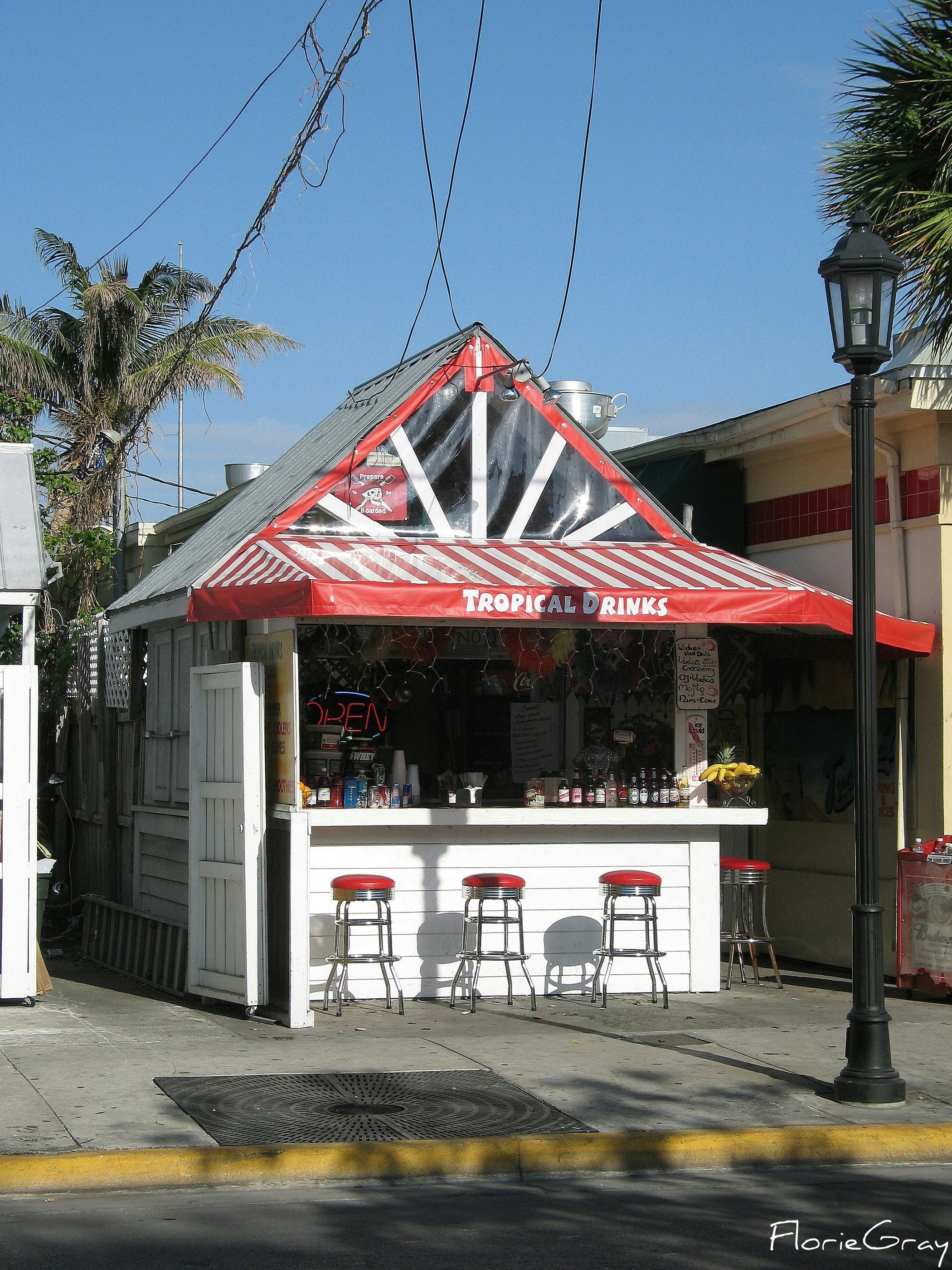 Photo captures a tropical drinks kiosk with a red-and-white striped roof and white counter on a sunlit street. Red stools line the front, and a tall street lamp stands nearby, adding urban contrast.