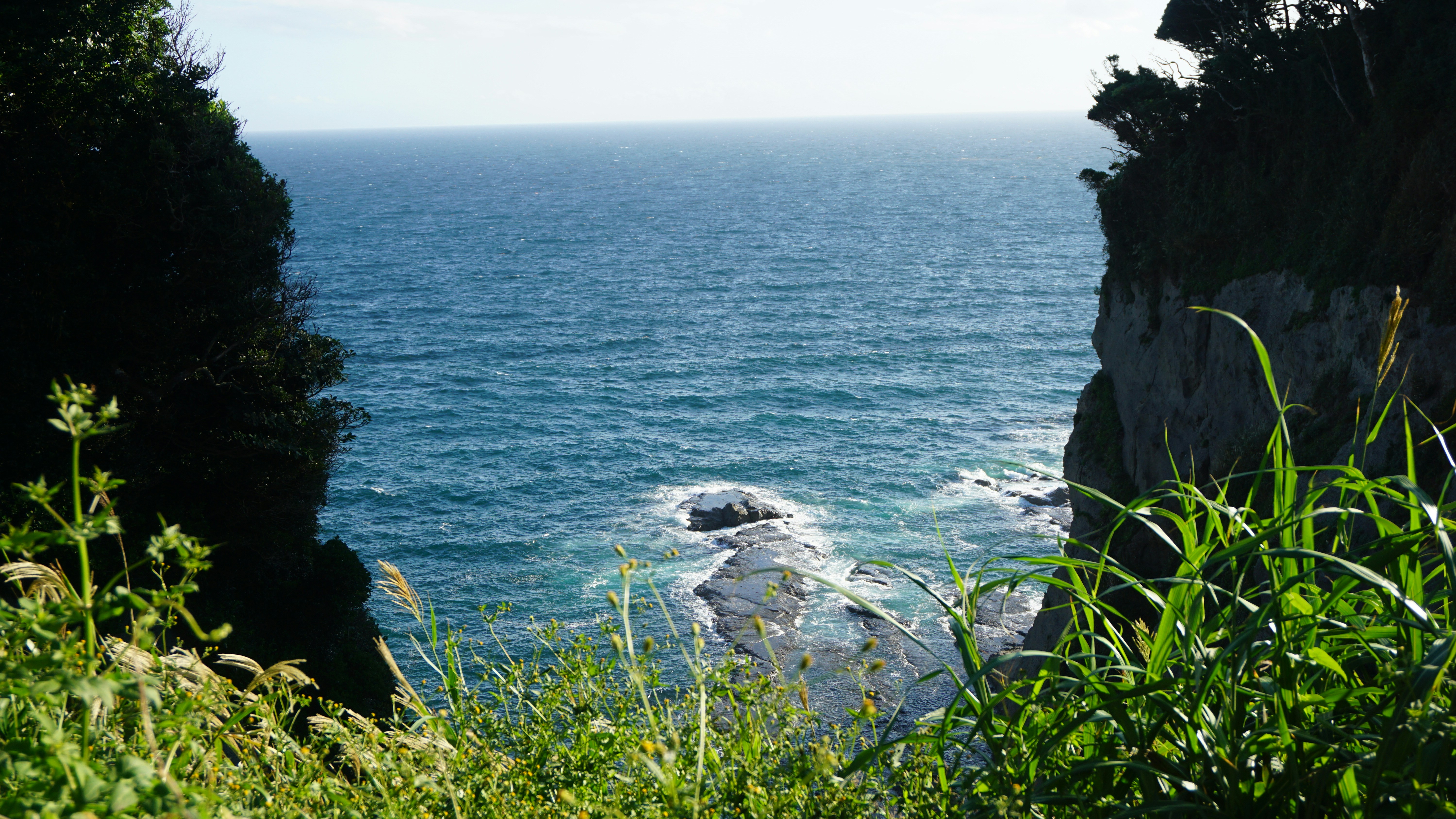Blue ocean waves viewed through a gap between lush green cliffs under a bright sky.