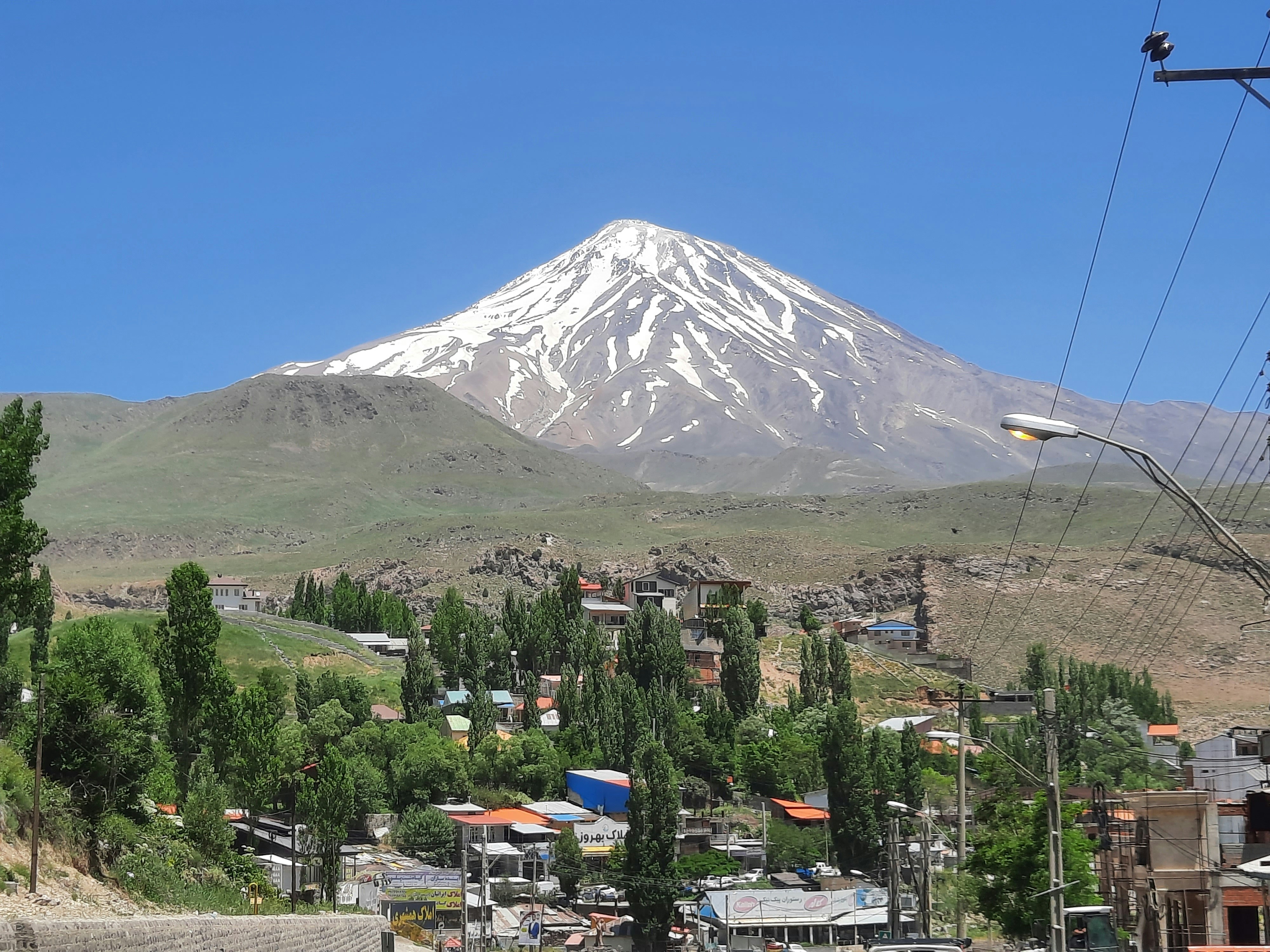 Snow-capped mountain towering over a lush valley dotted with colorful homes and trees. A clear blue sky enhances the vibrant landscape.