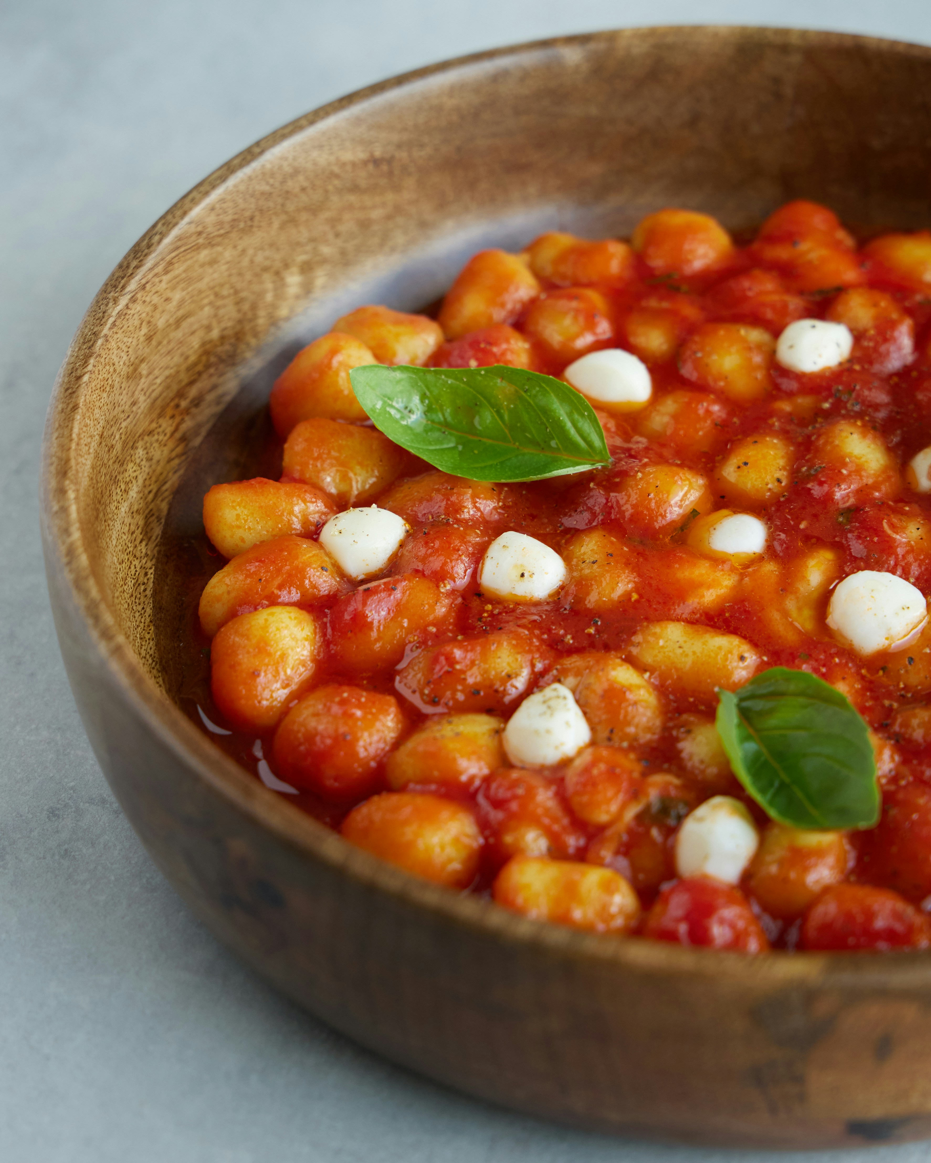 A wooden bowl filled with gnocchi in rich tomato sauce, topped with fresh basil and mozzarella pearls.