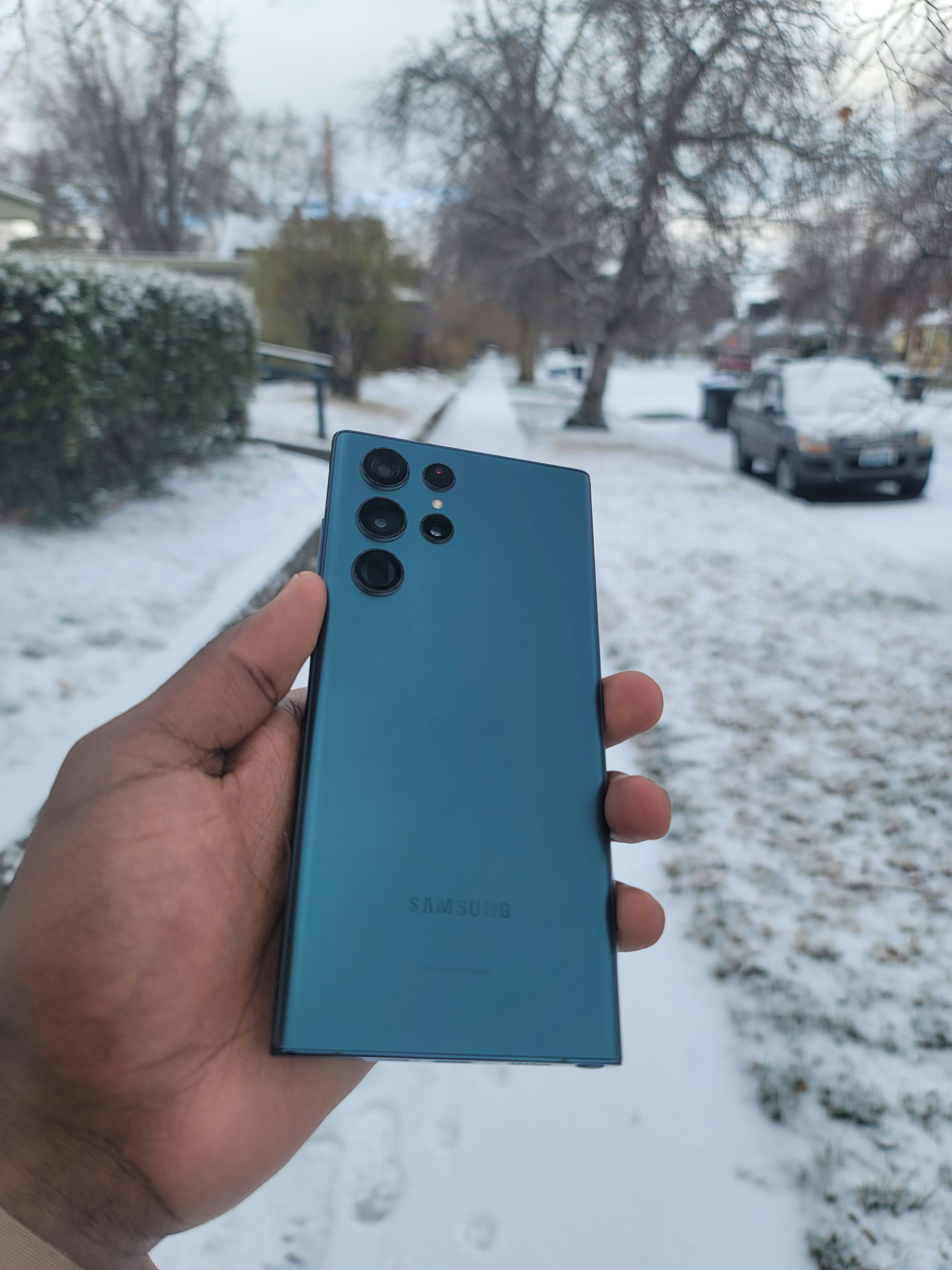 A teal Samsung Galaxy smartphone with a multi-lens rear camera is held in a dark-skinned hand, with a snow-covered suburban street in the background.