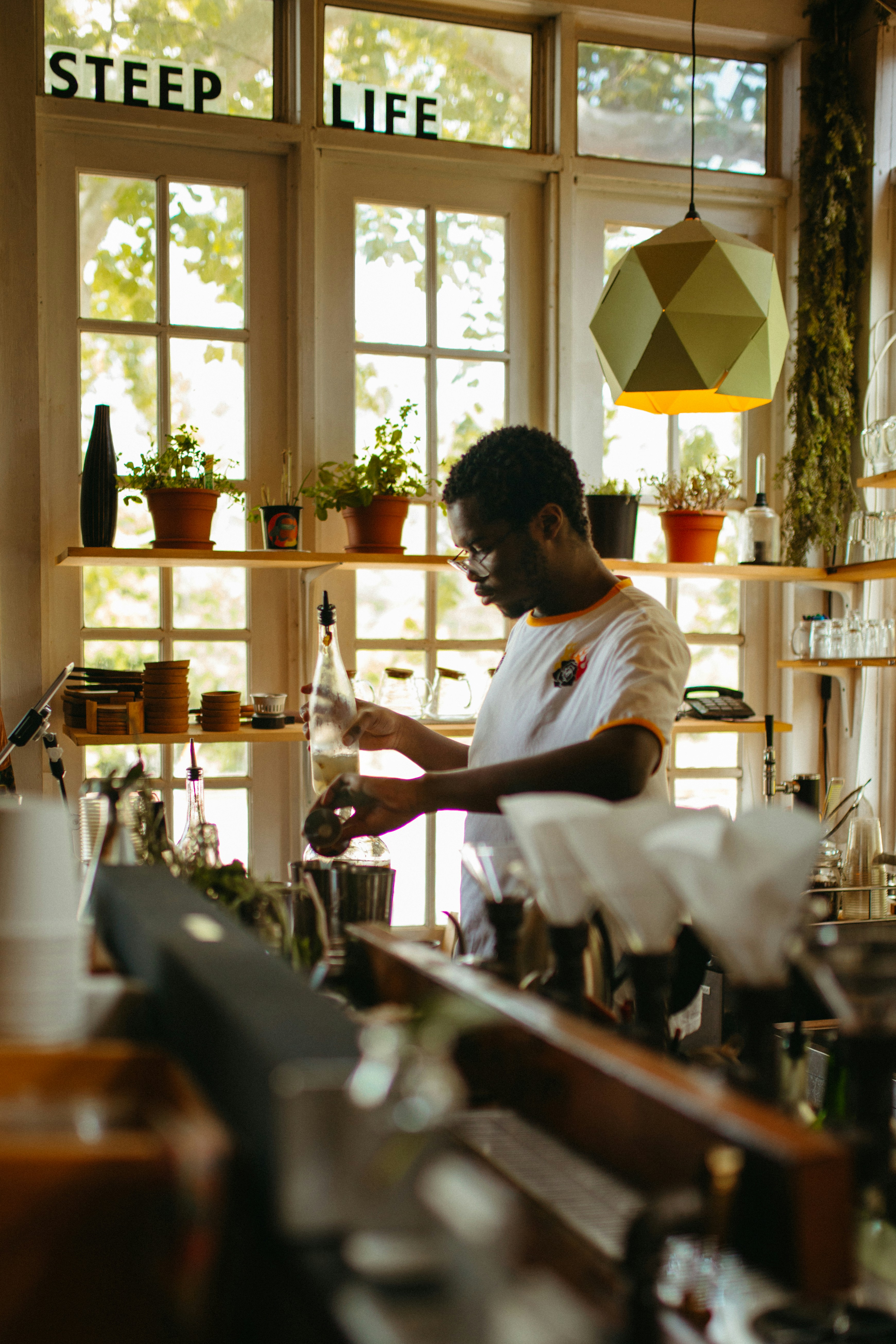 Sunlit Barista at a Plant-Lined Counter Crafts a Quiet Pour
