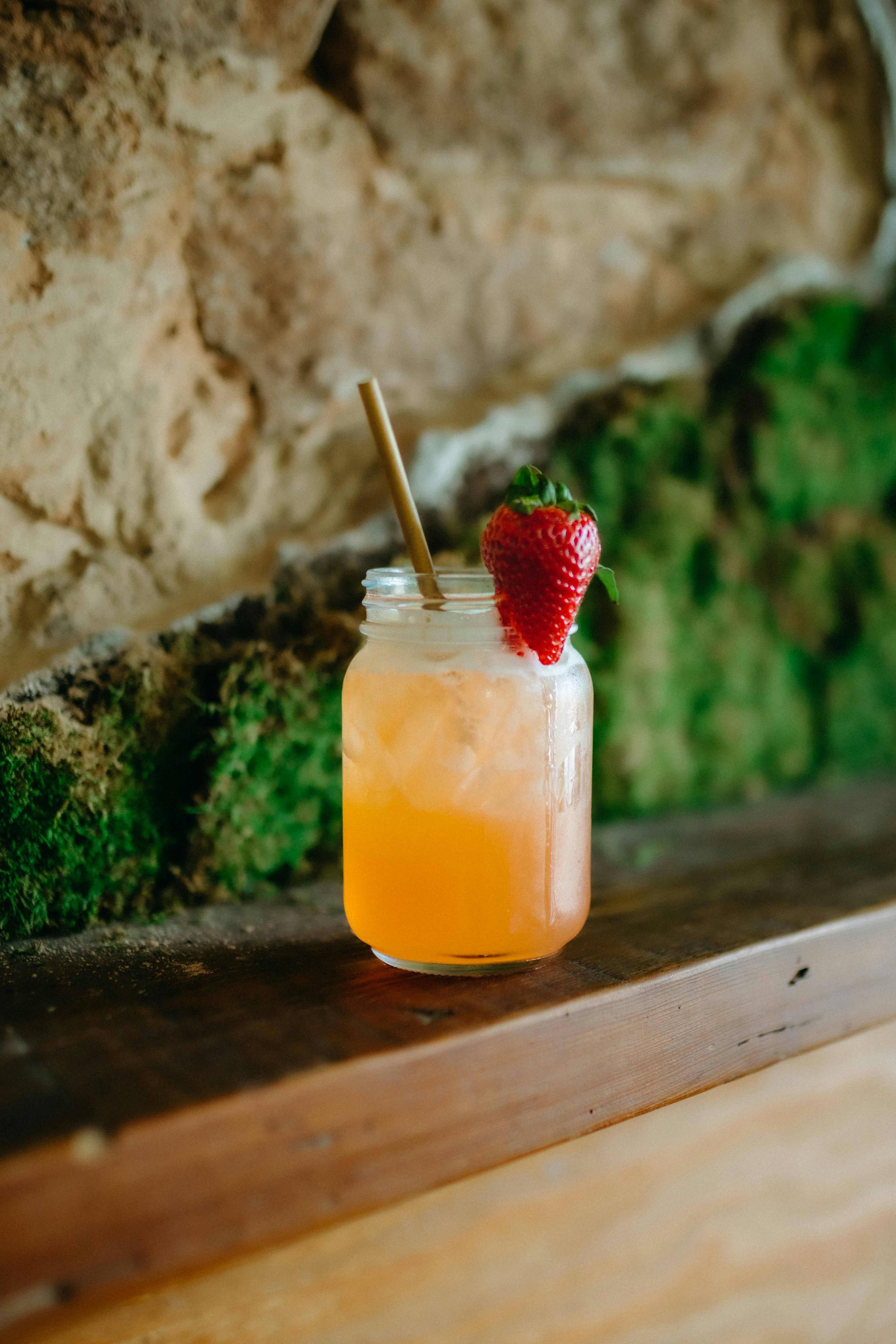 A mason jar filled with a vibrant orange drink, garnished with a fresh strawberry, sits on a wooden shelf against a textured stone wall and lush green moss.