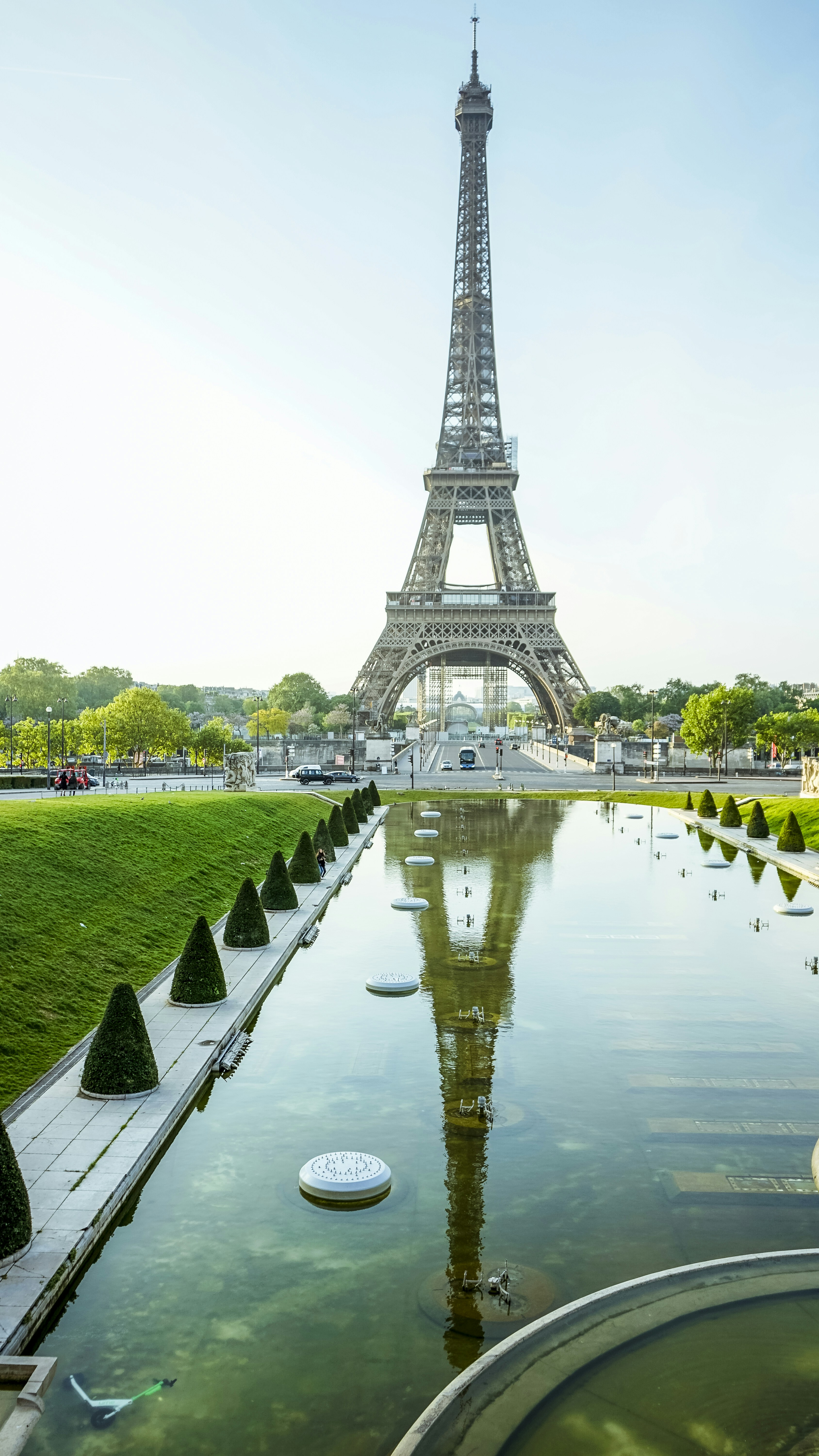 Eiffel Tower rises above a long reflecting pool bordered by neatly trimmed conical topiaries in a sunlit Paris garden.