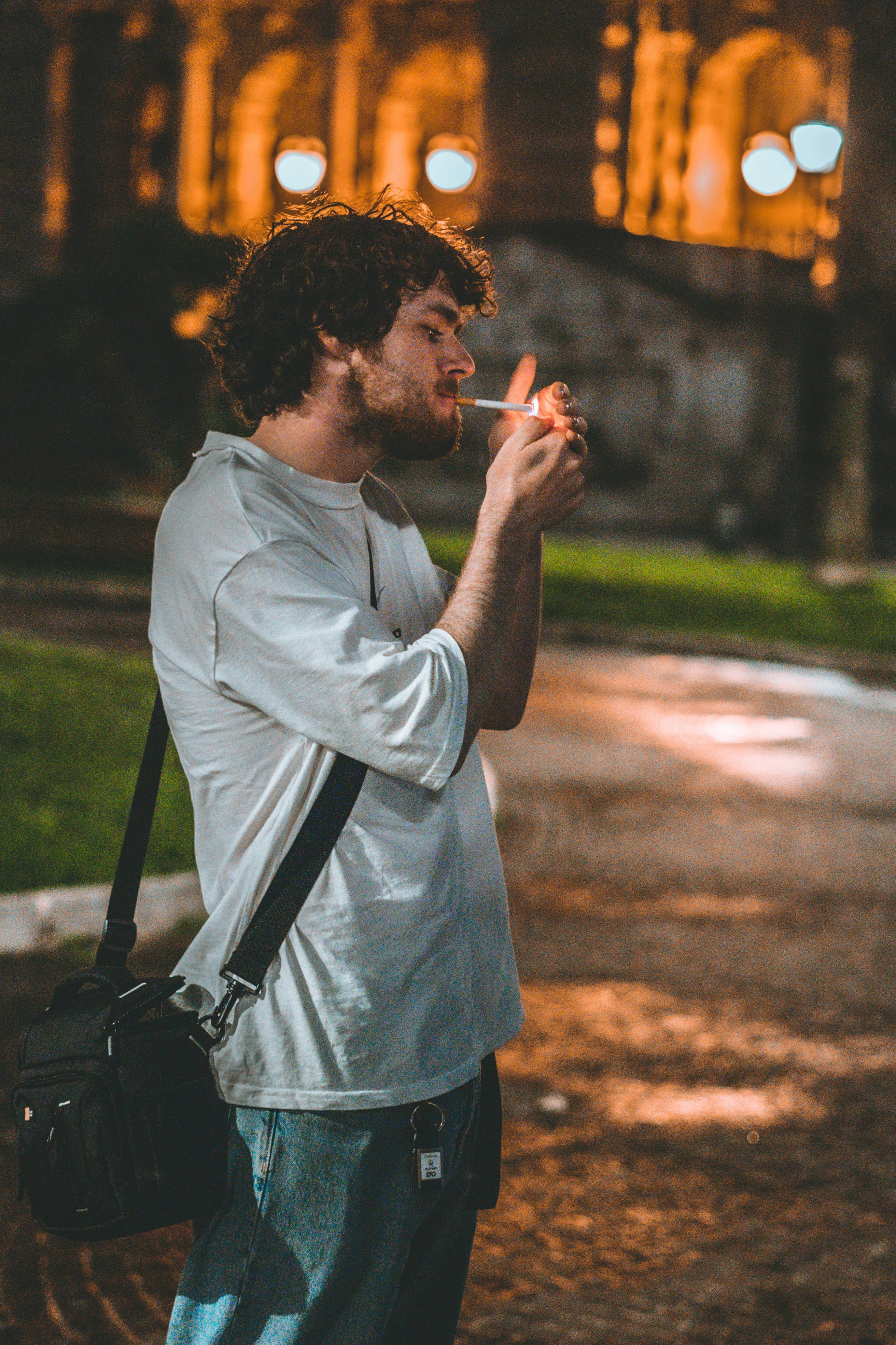 A young man stands in a softly lit park, lighting a cigarette while lost in thought. The warm glow of nearby lamps creates a serene atmosphere.