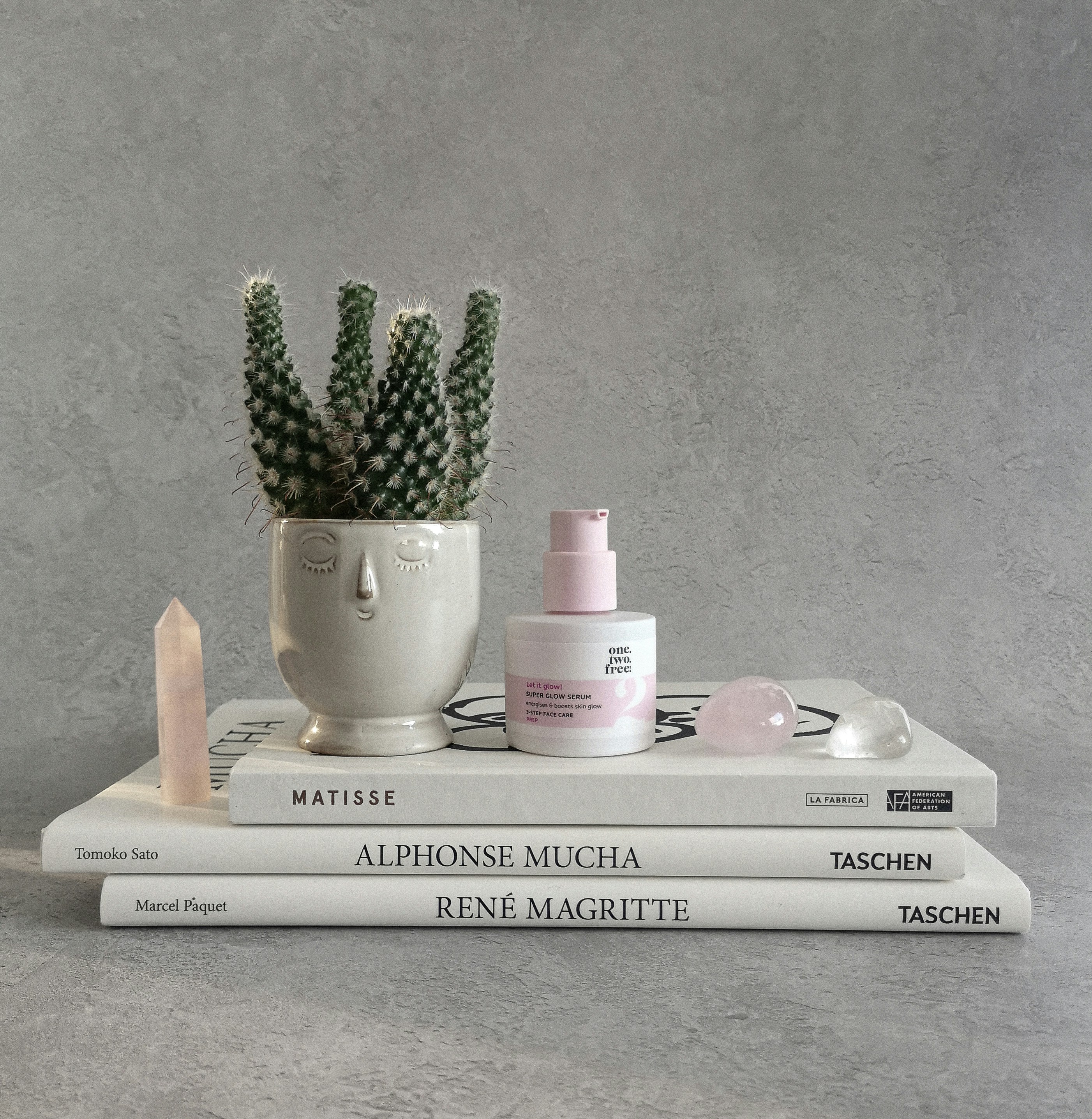A minimalist arrangement featuring a potted cactus, skincare product, and various crystals atop art books. The soft gray background enhances the serene aesthetic.