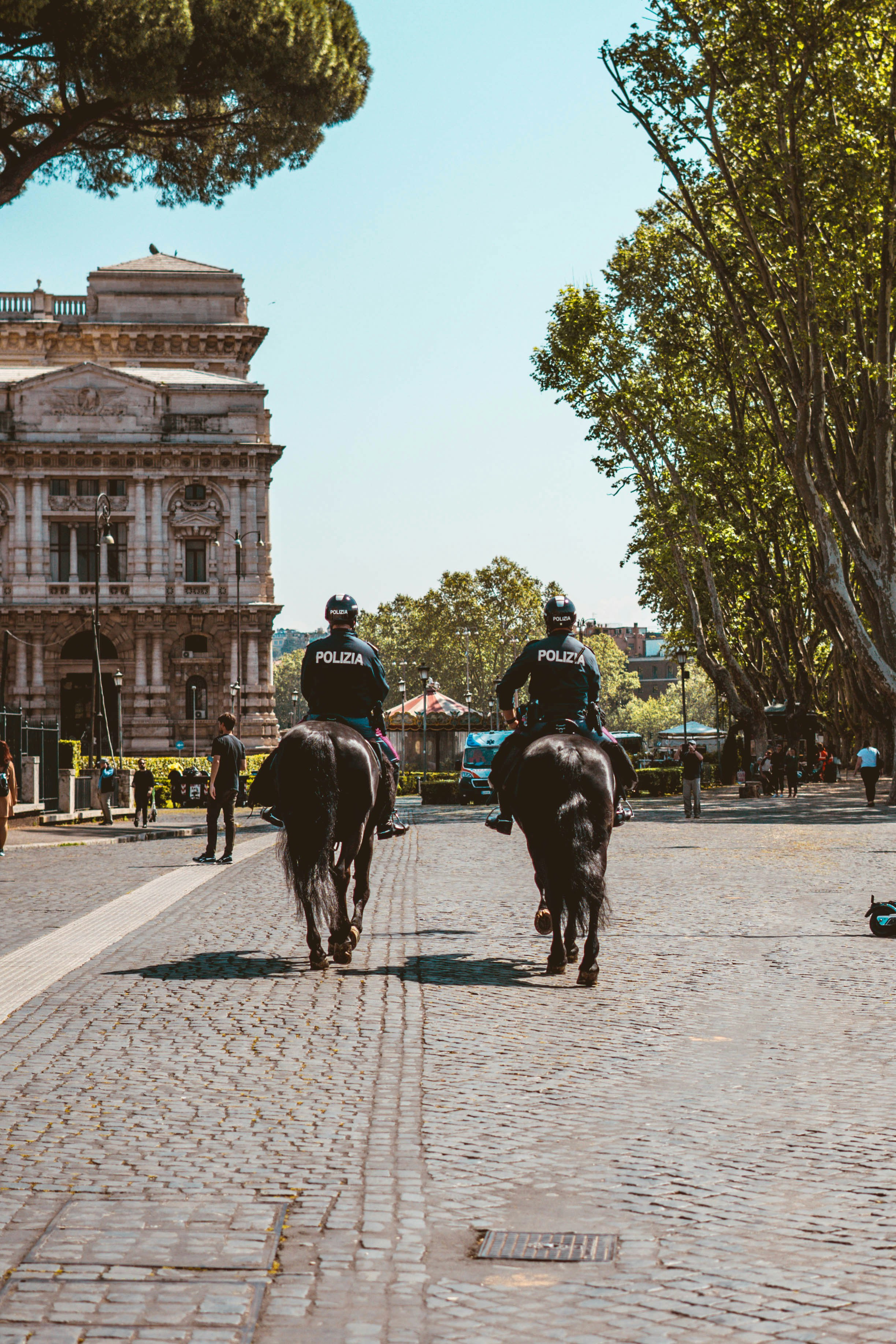 Two mounted police officers patrol a cobblestone street lined with trees and historic architecture. The scene captures a blend of urban life and law enforcement.