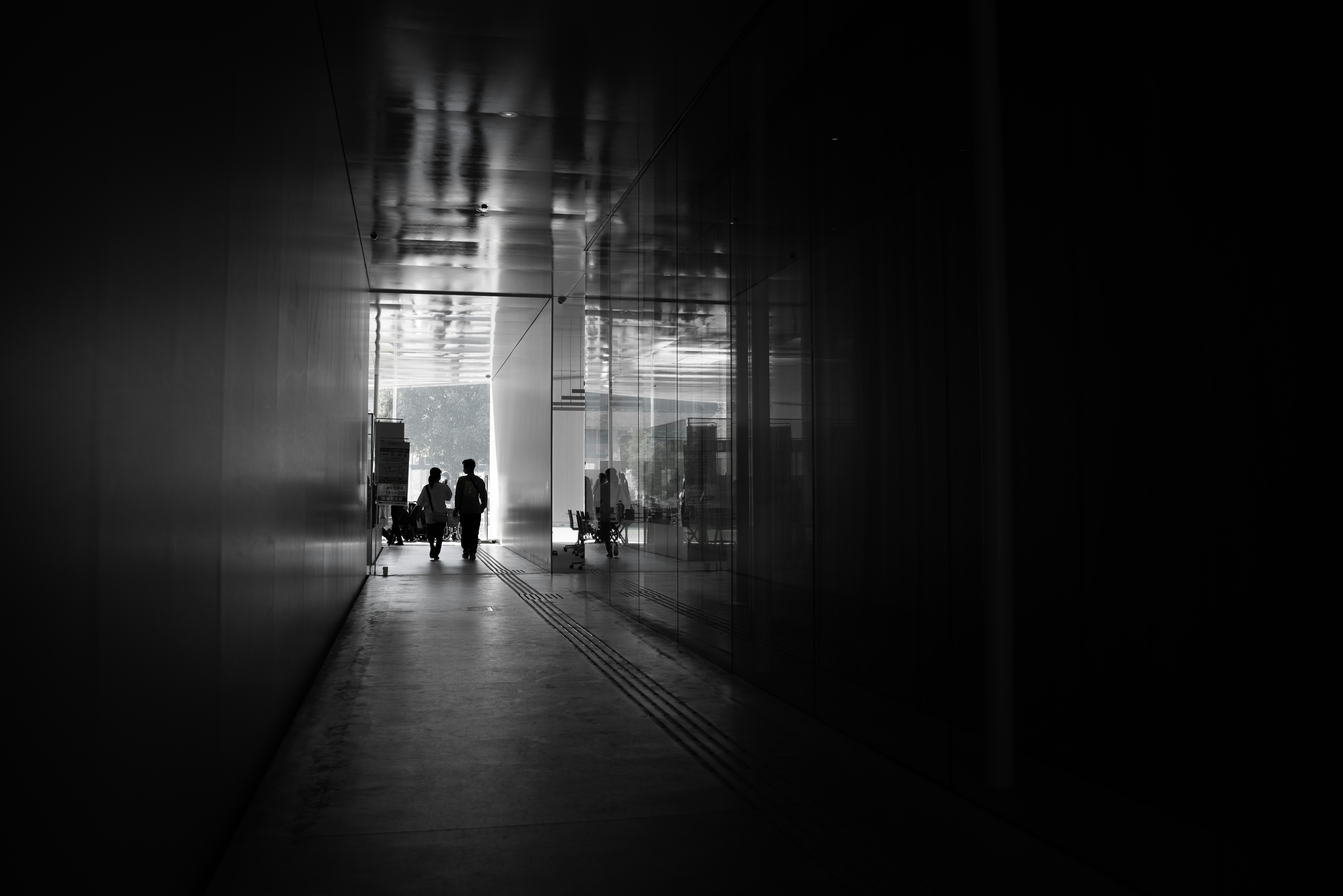 Silhouetted figures walk through a dimly lit corridor, illuminated by a bright exit at the end, creating a striking contrast between light and shadow.