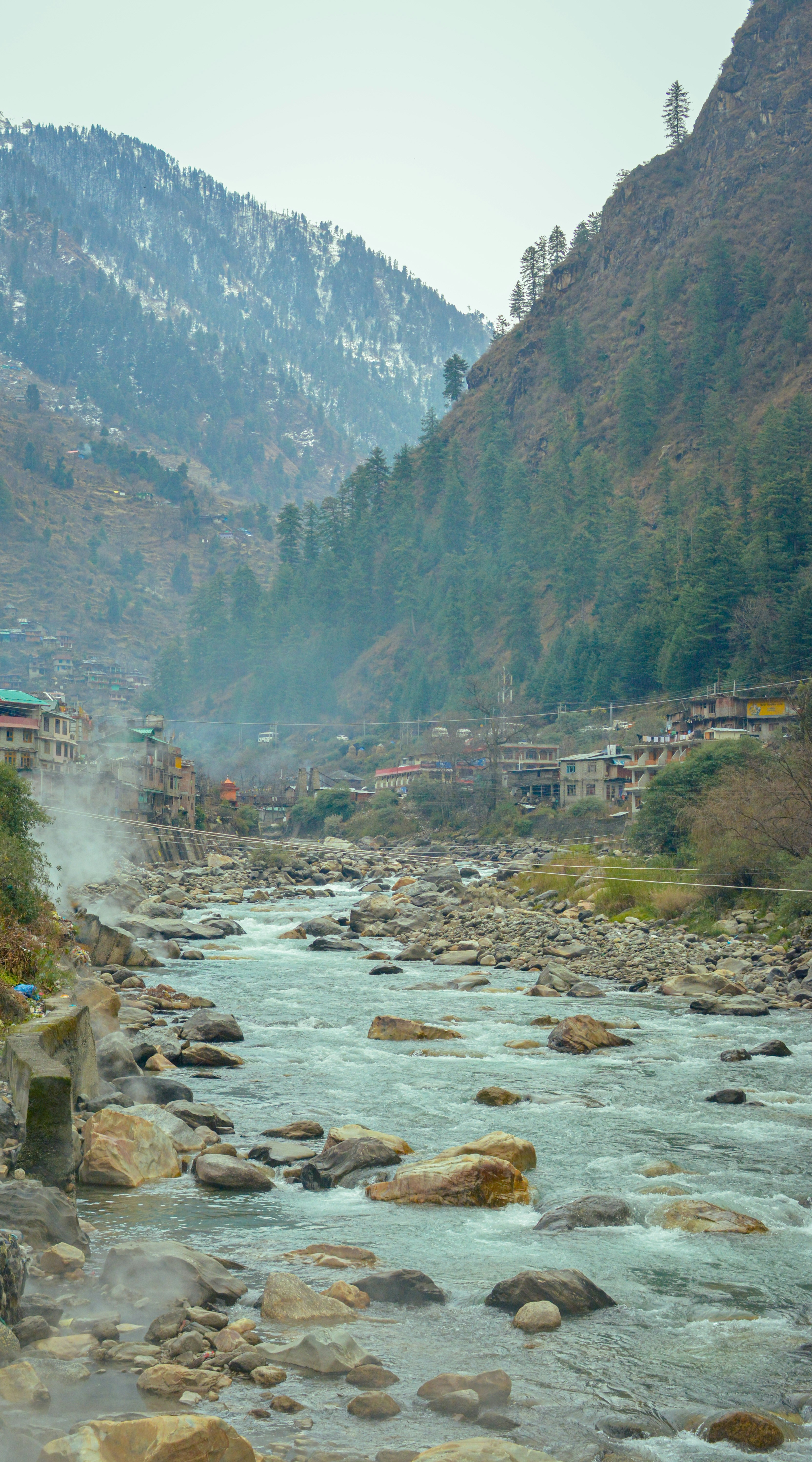 Flowing river bordered by rocky banks and quaint buildings nestled in the mountains, shrouded in mist. A tranquil scene capturing the essence of nature and human coexistence.