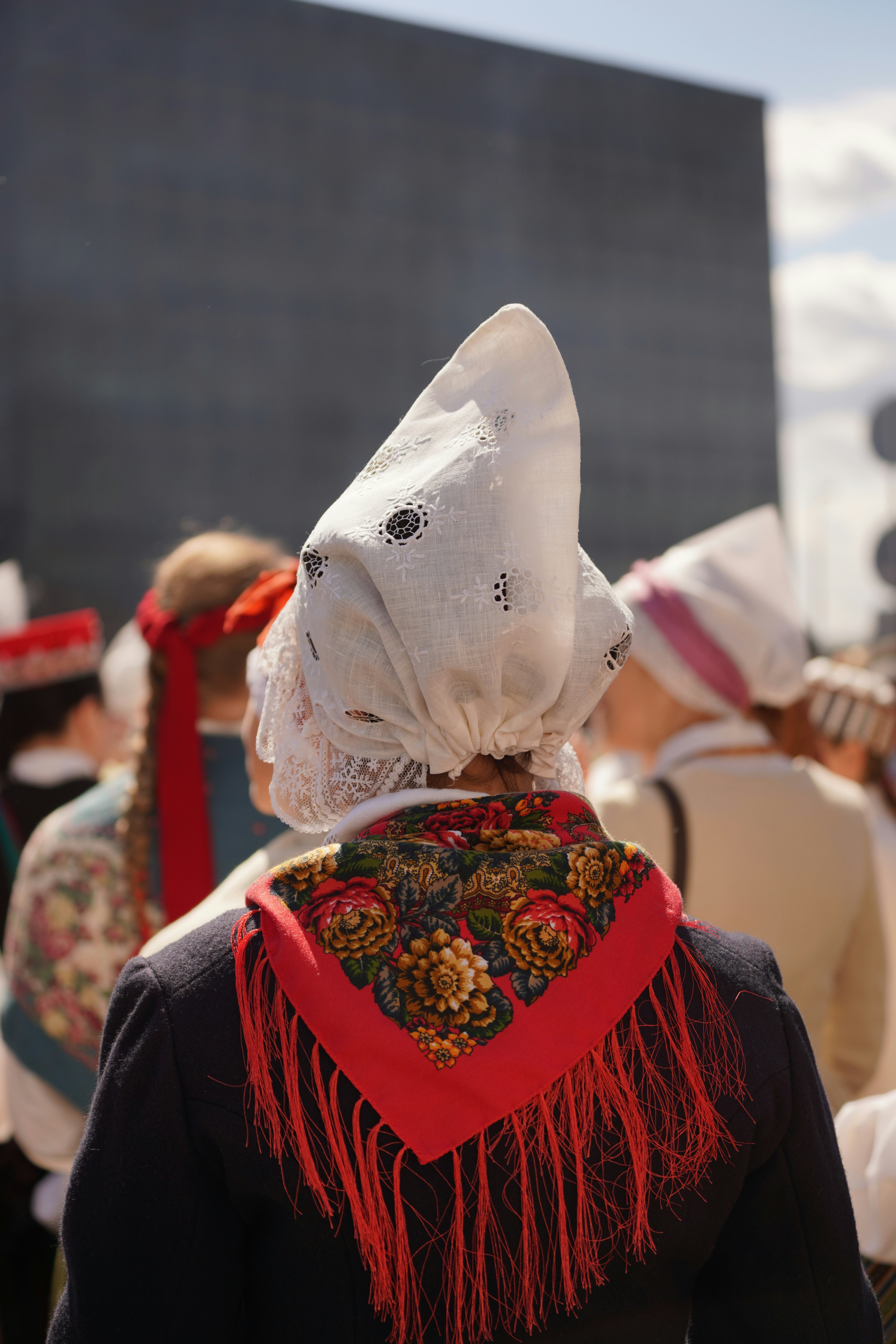 A woman in traditional attire, adorned with a vibrant floral scarf and a lace head covering, stands amidst a gathering, embodying cultural heritage.