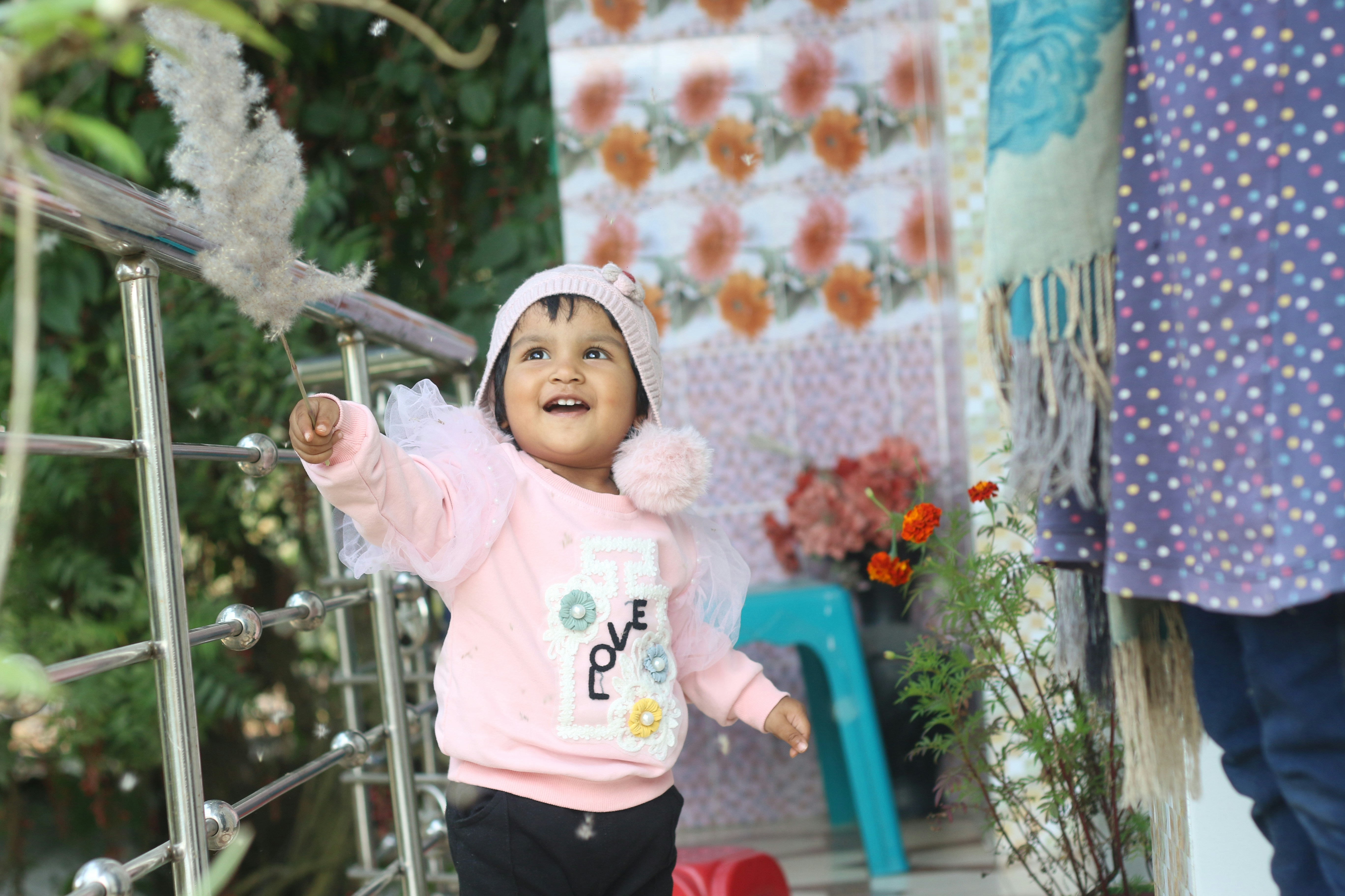 Smiling toddler in a pink outfit reaching out on a decorated balcony with flowers and patterned backdrop.