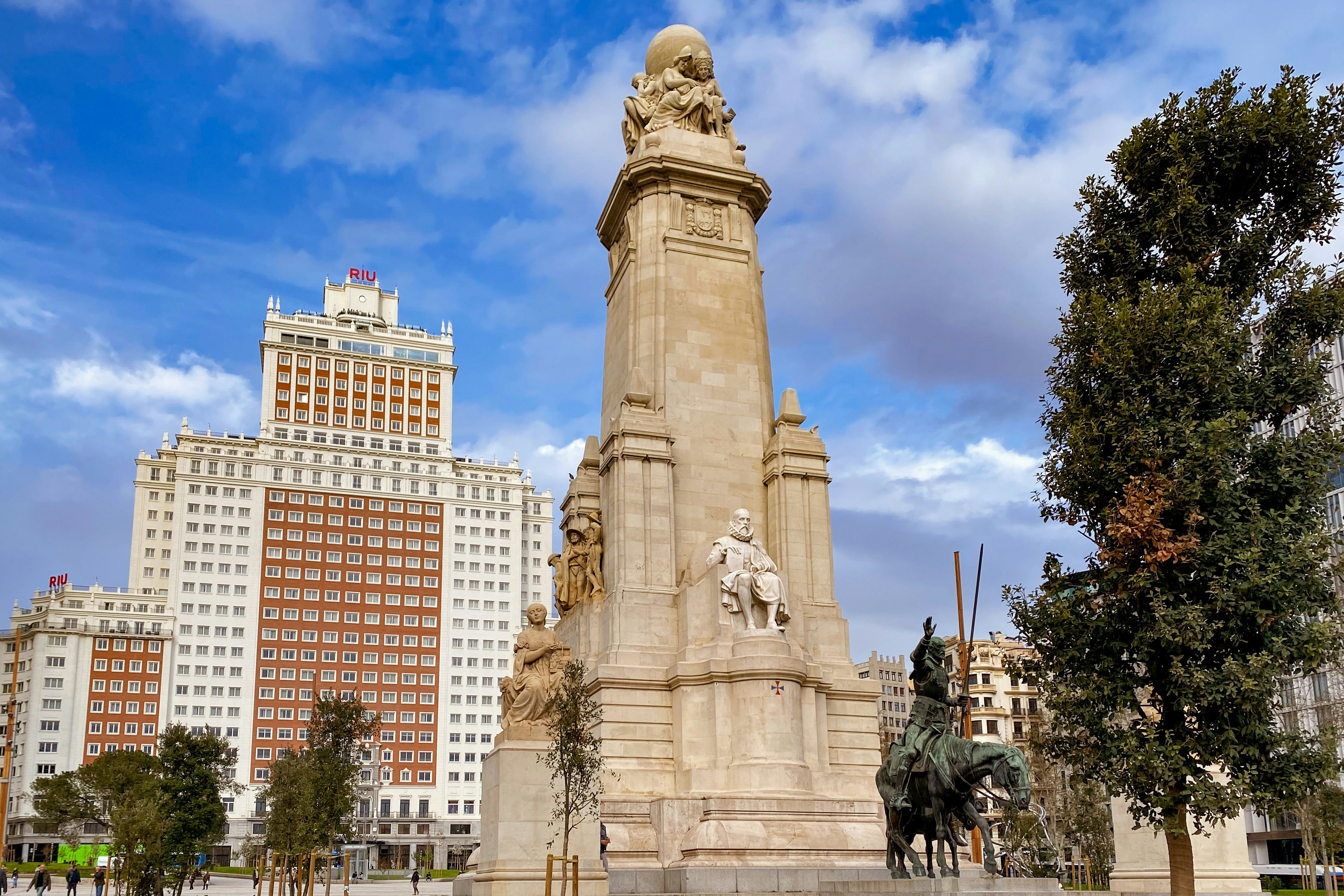 A grand monument featuring historical figures, surrounded by modern architecture and greenery. The scene captures the blend of past and present in an urban setting.