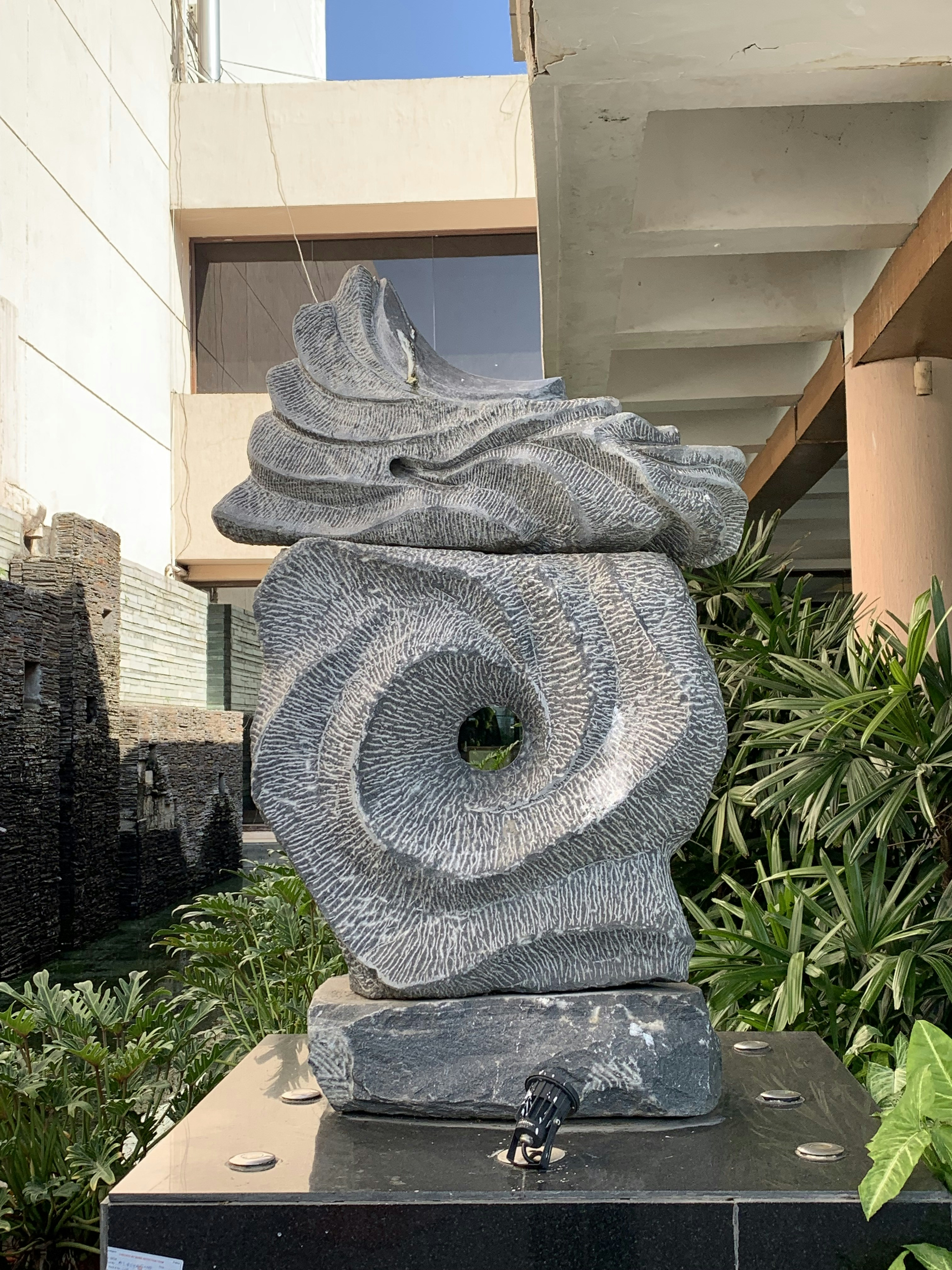 Abstract stone sculpture featuring swirling patterns and a central void, surrounded by lush greenery and modern architecture.