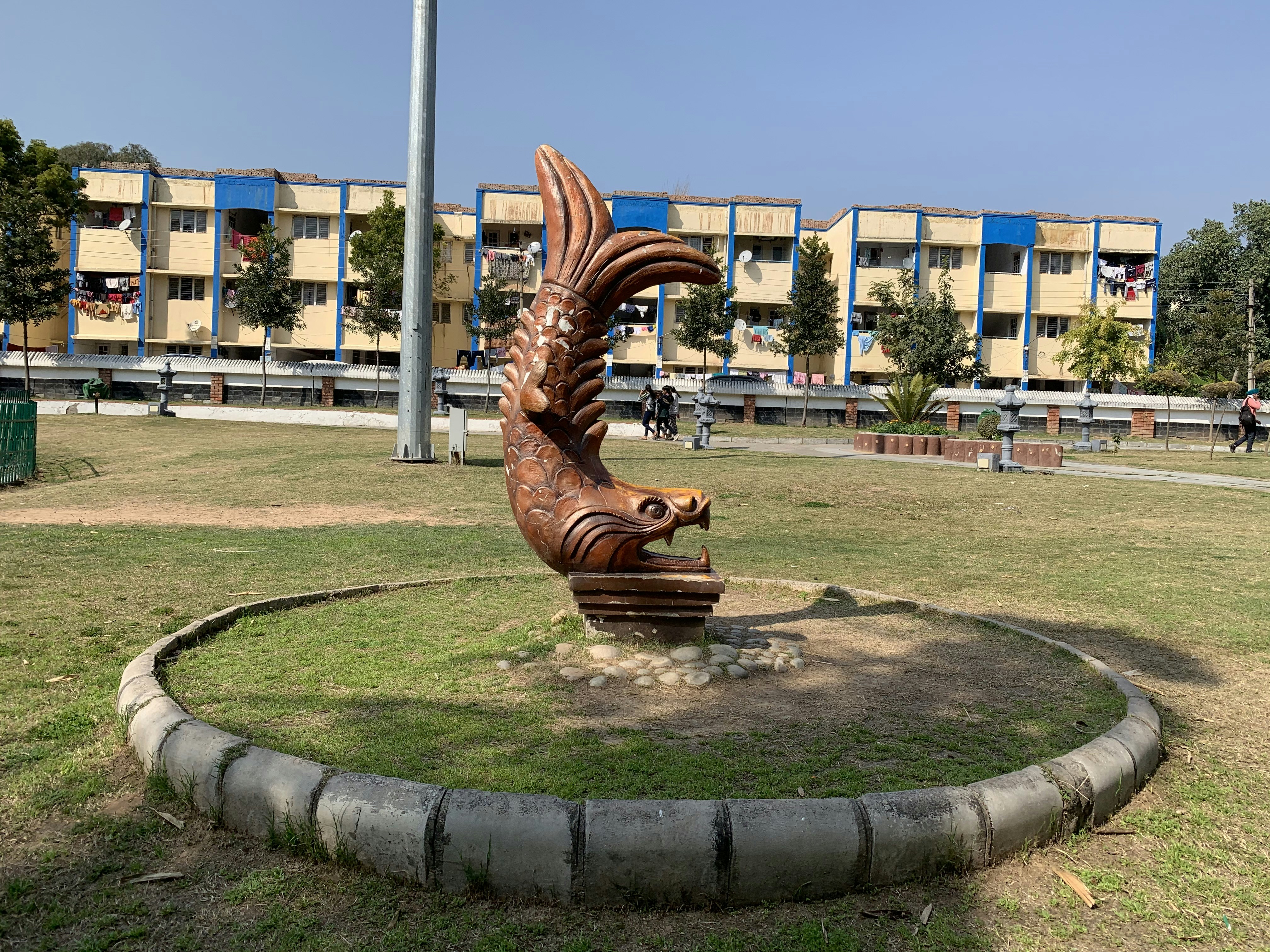Intricately carved fish sculpture surrounded by a circular stone base in a grassy park area, with residential buildings in the background.