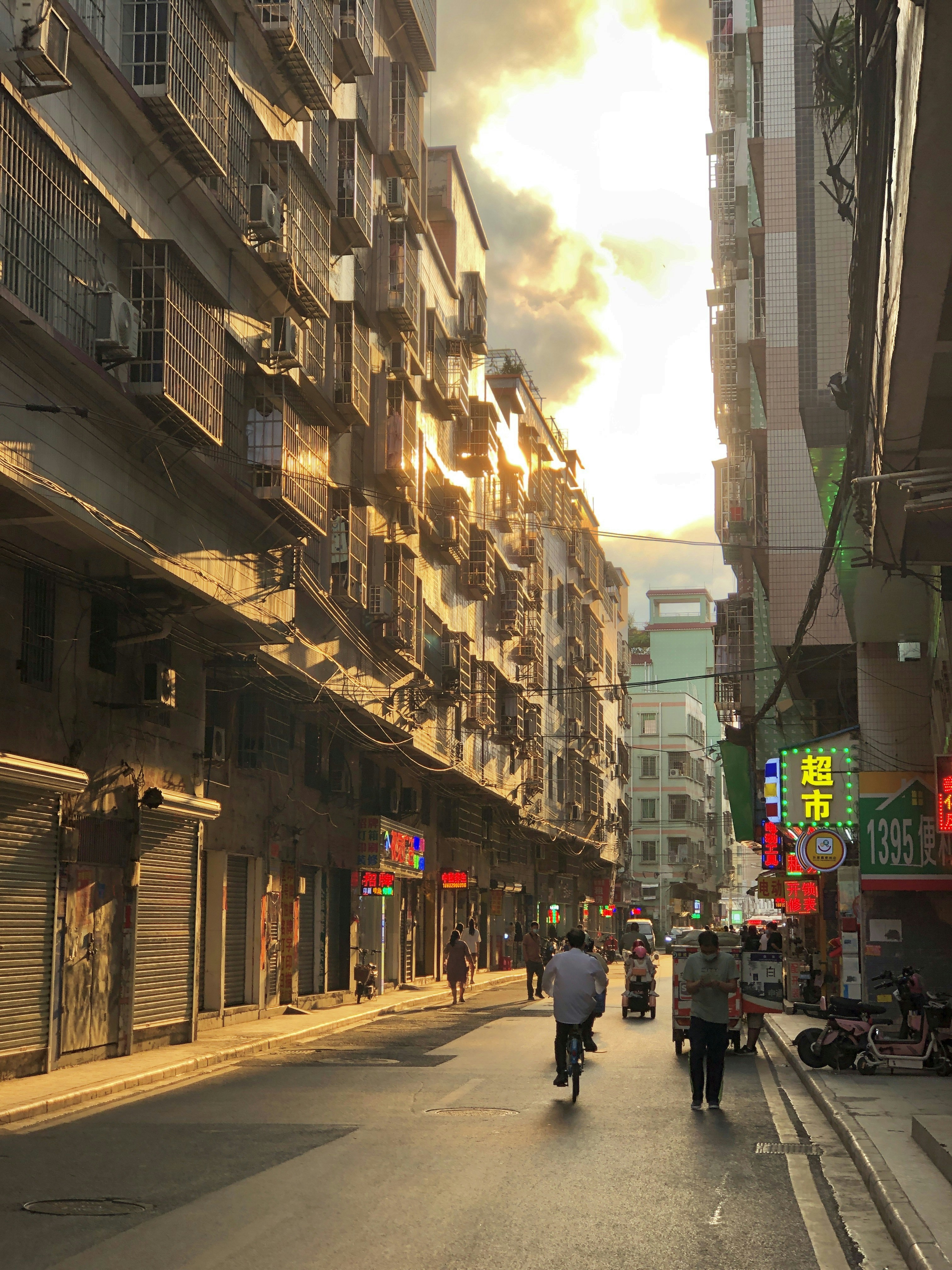 Narrow city street illuminated by warm sunset light, showcasing buildings adorned with air conditioning units and vibrant neon signs. A cyclist navigates the scene.