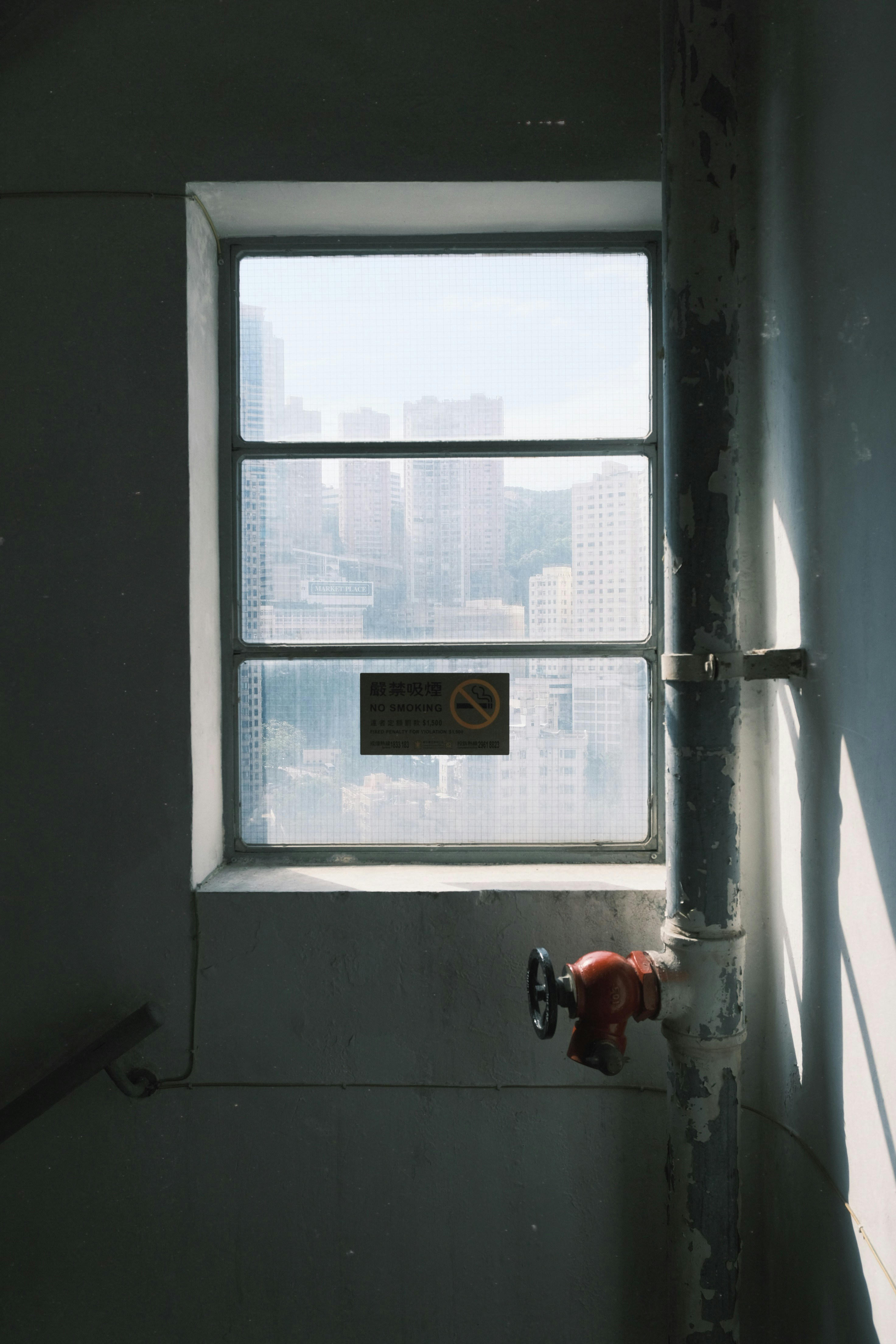 Industrial window revealing a cityscape with towering buildings and a warning sign. A red valve adds a touch of character to the scene.