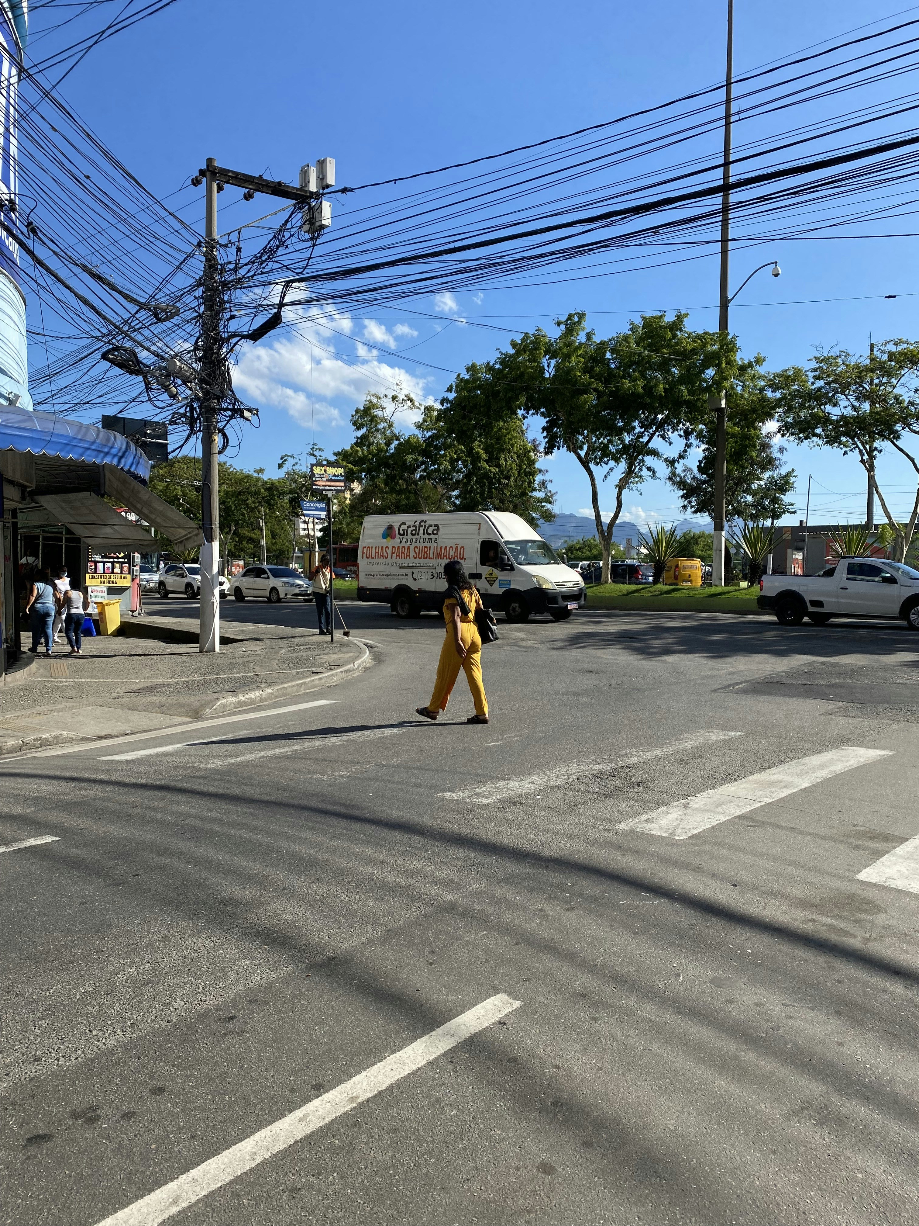 A pedestrian in yellow pants crosses a bustling street, framed by urban elements and vibrant greenery. The scene captures the essence of city life.