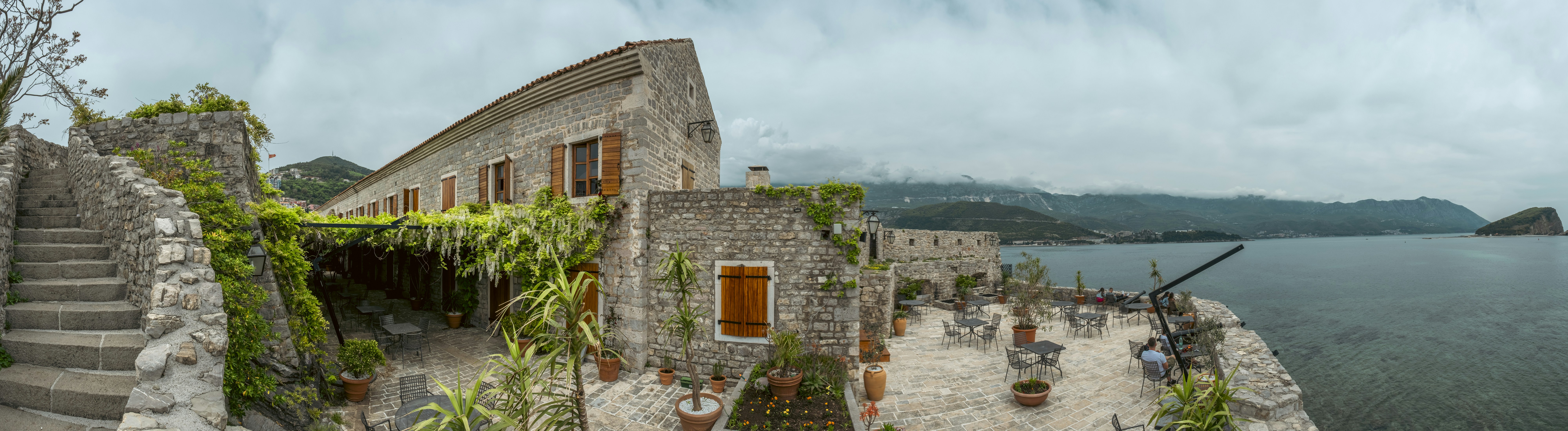 A panoramic view of a historic stone building adorned with greenery, overlooking a serene body of water under a cloudy sky.
