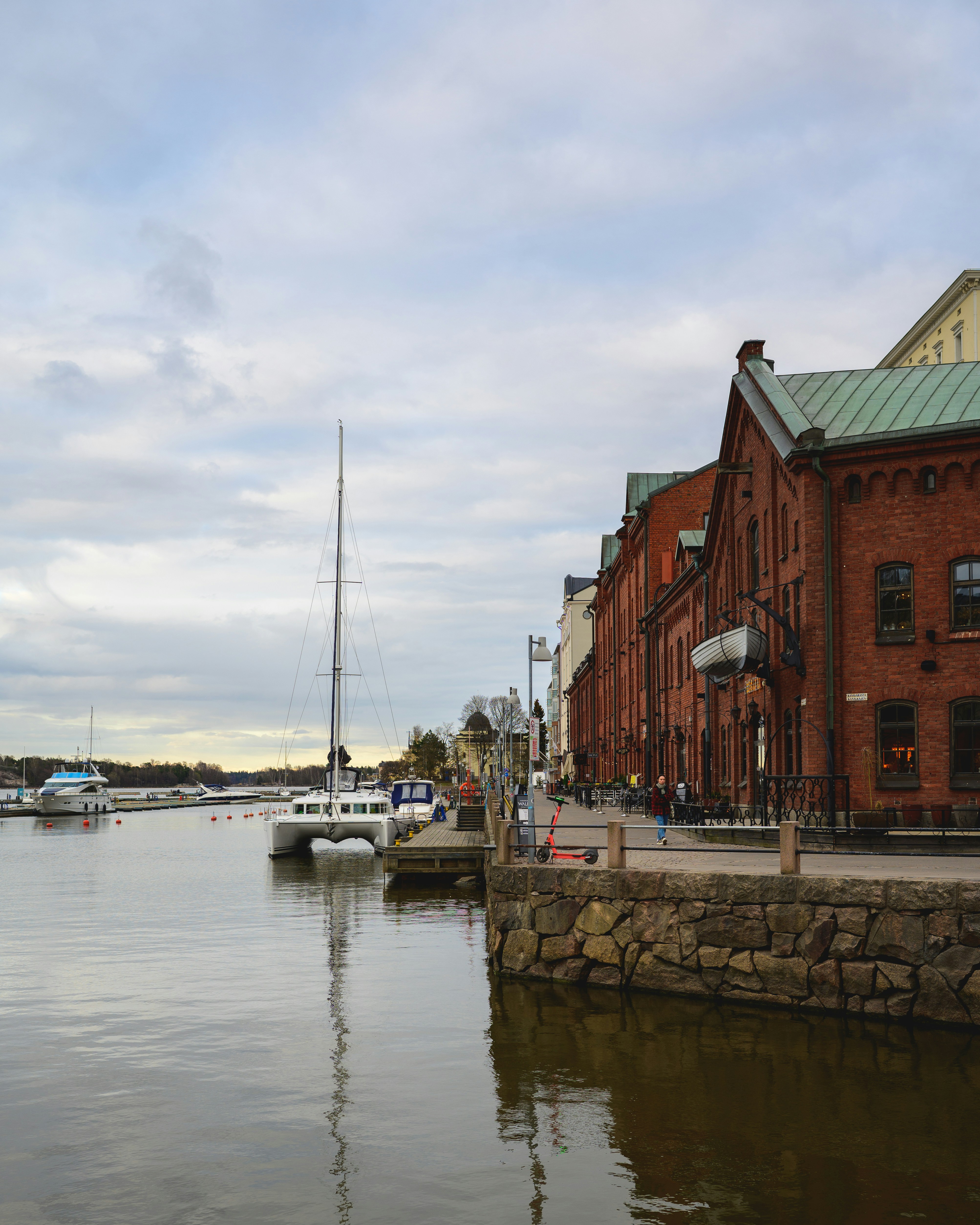 Historic red brick building alongside a serene harbor, with sailboats and a cloudy sky reflecting on the water's surface.