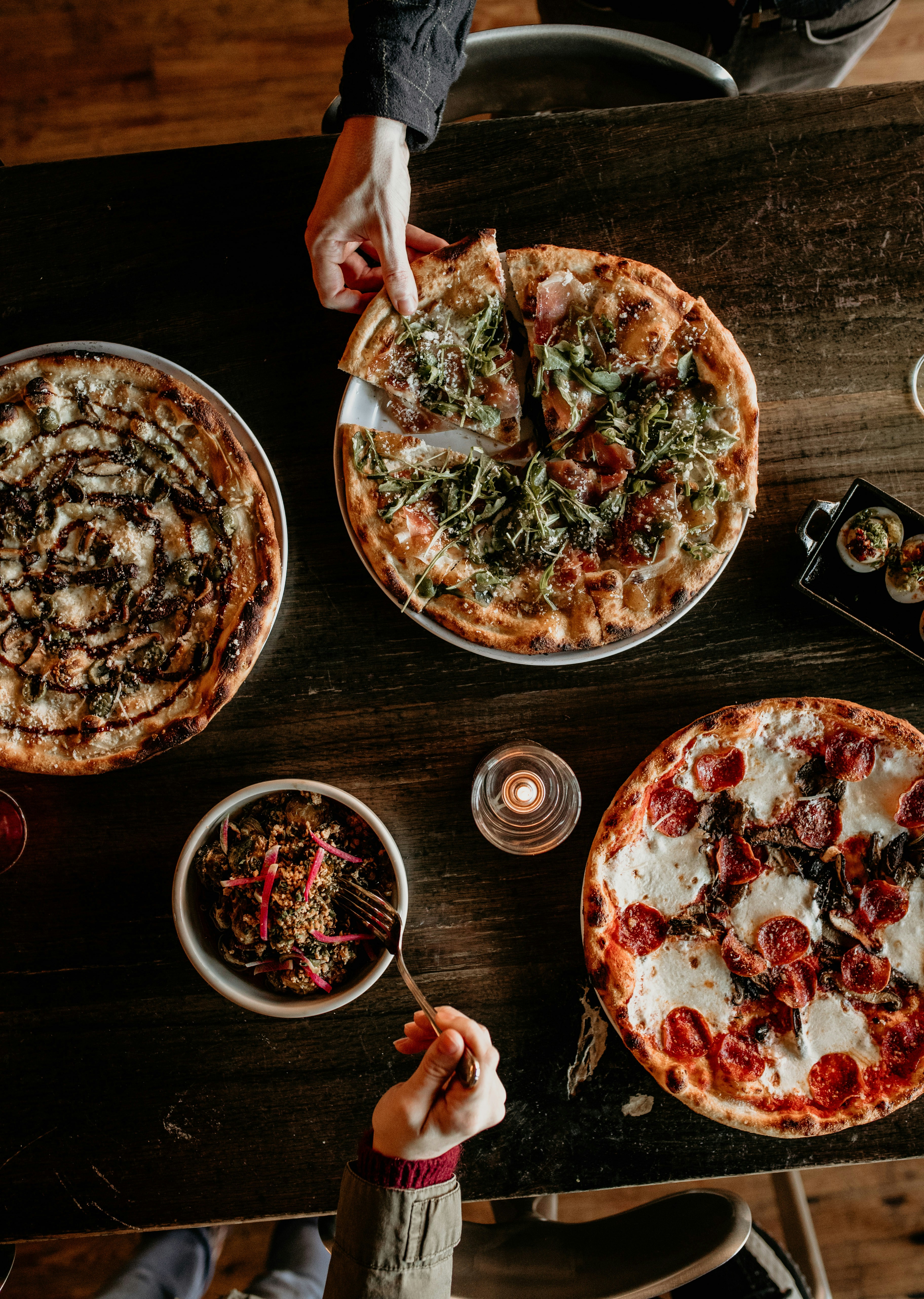 Three gourmet pizzas and a colorful salad on a rustic wooden table, with hands reaching for the food. A candle flickers nearby, adding warmth to the scene.