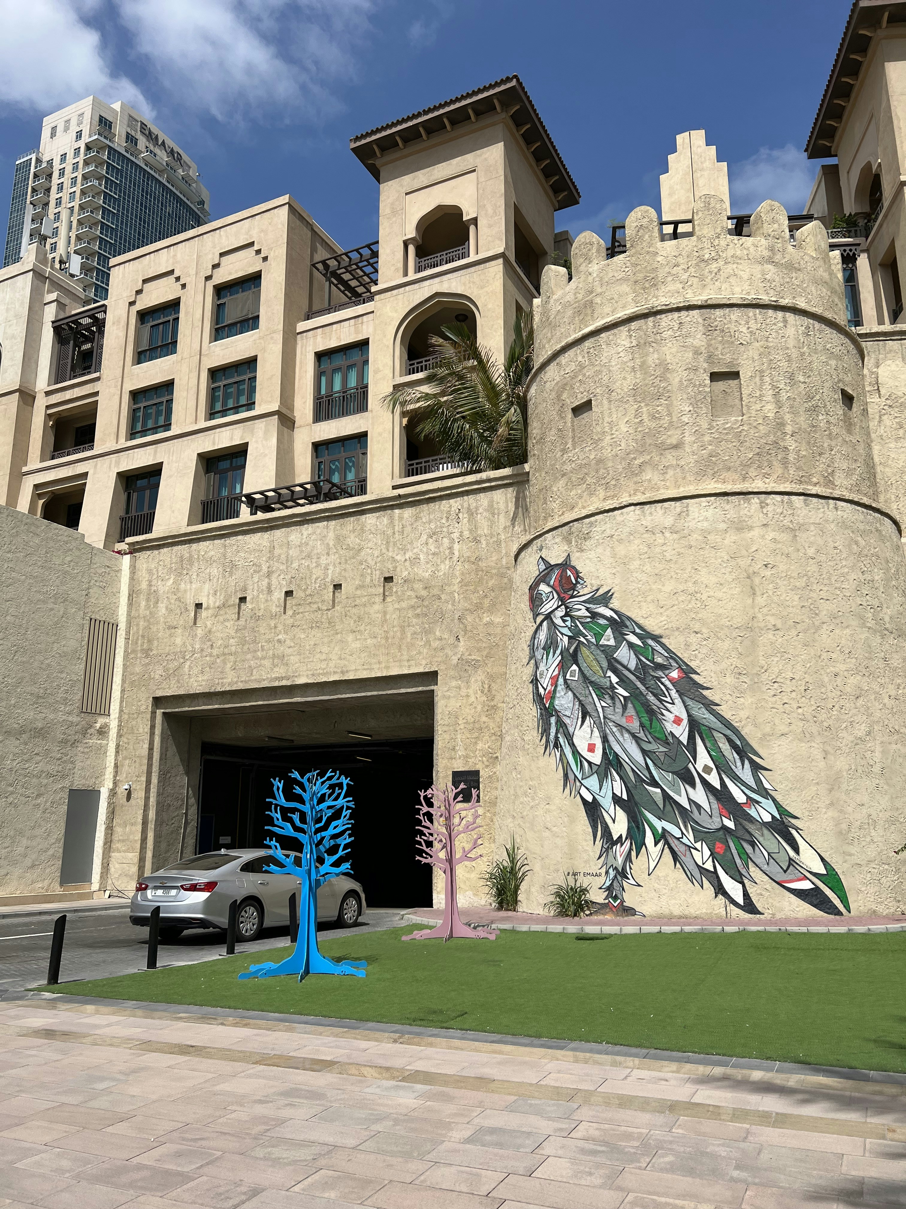 A vibrant mural of a bird adorns a textured wall beside modern architecture, with playful, colorful trees in the foreground. The scene captures the essence of urban creativity.