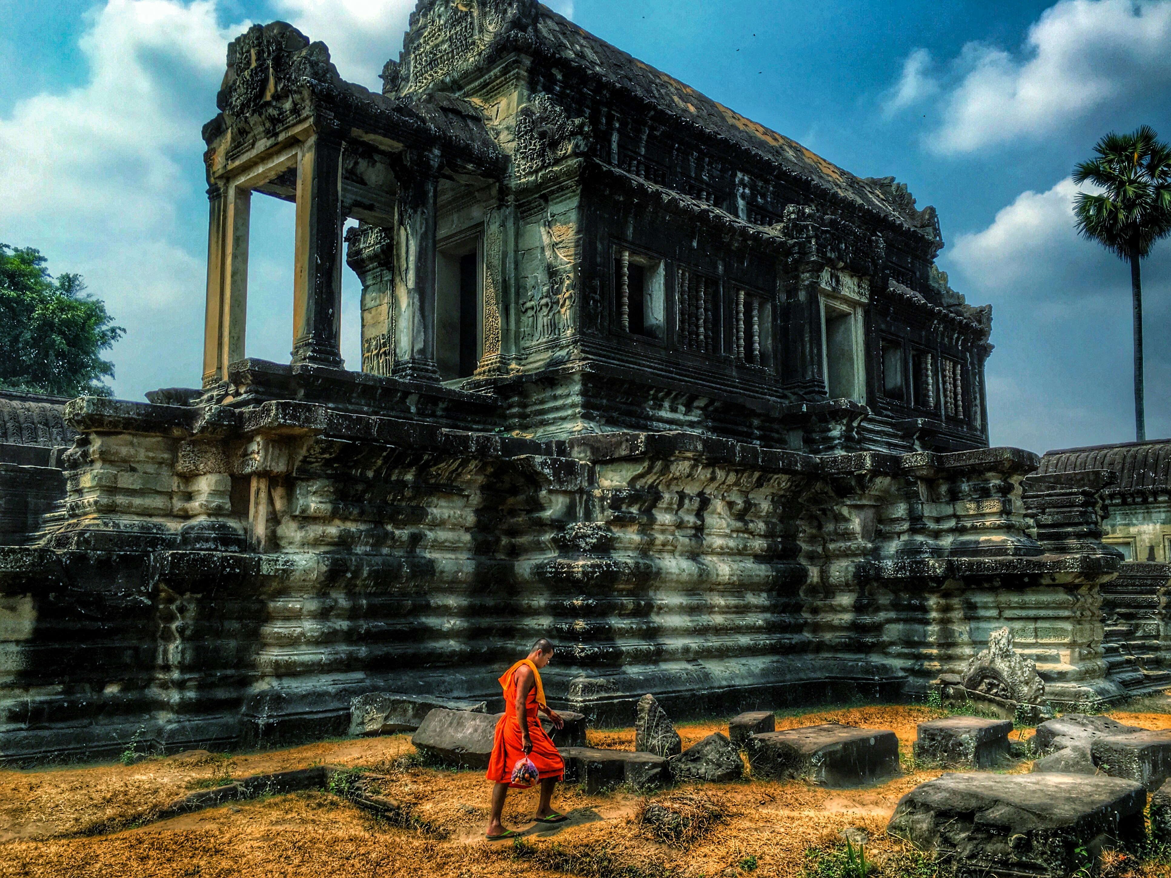 A monk in a vibrant orange robe walks among the weathered stones of an ancient temple, surrounded by lush greenery and a bright blue sky.