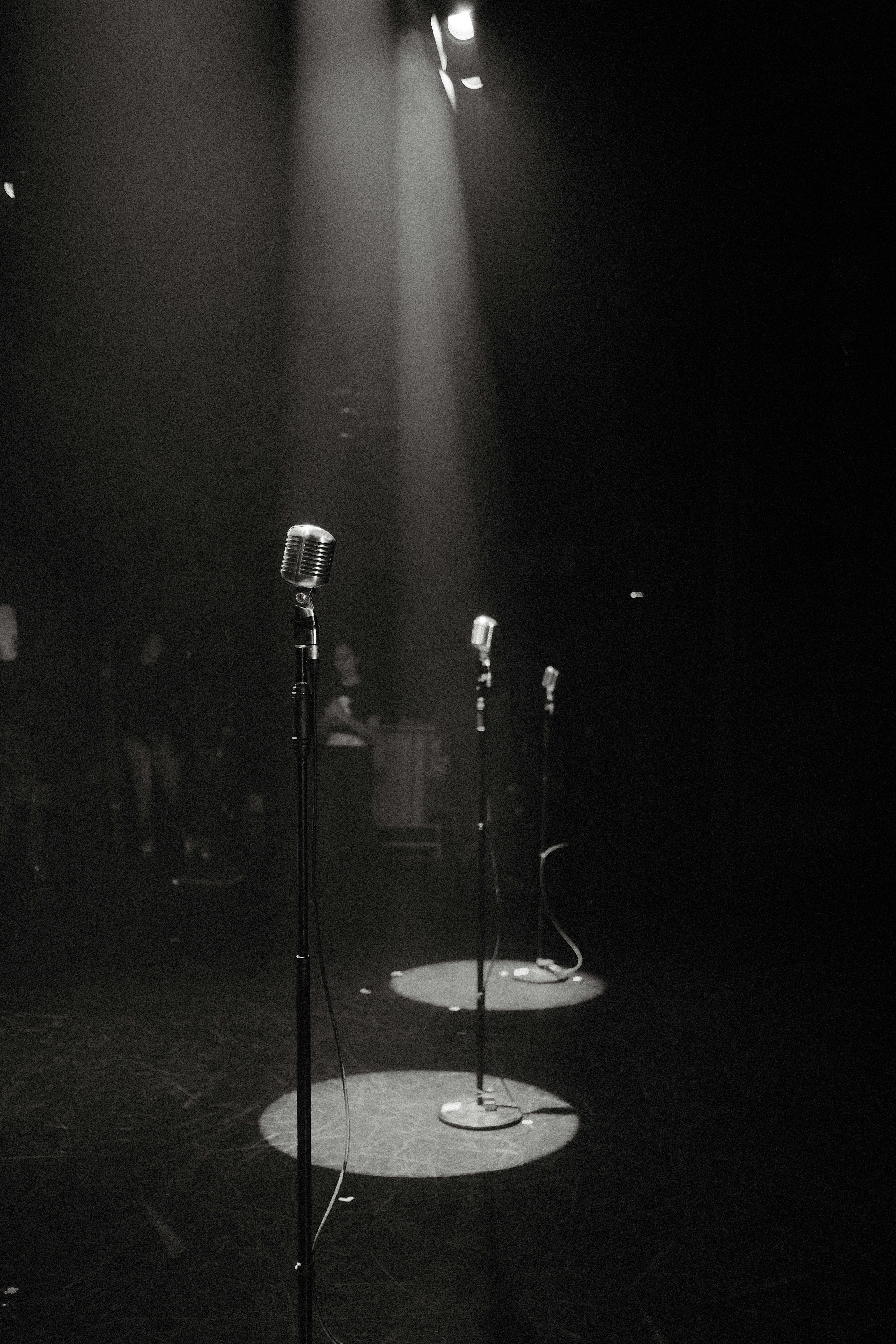Three vintage microphones stand under focused beams of light, casting circular shadows on the stage, evoking a sense of readiness for performance.