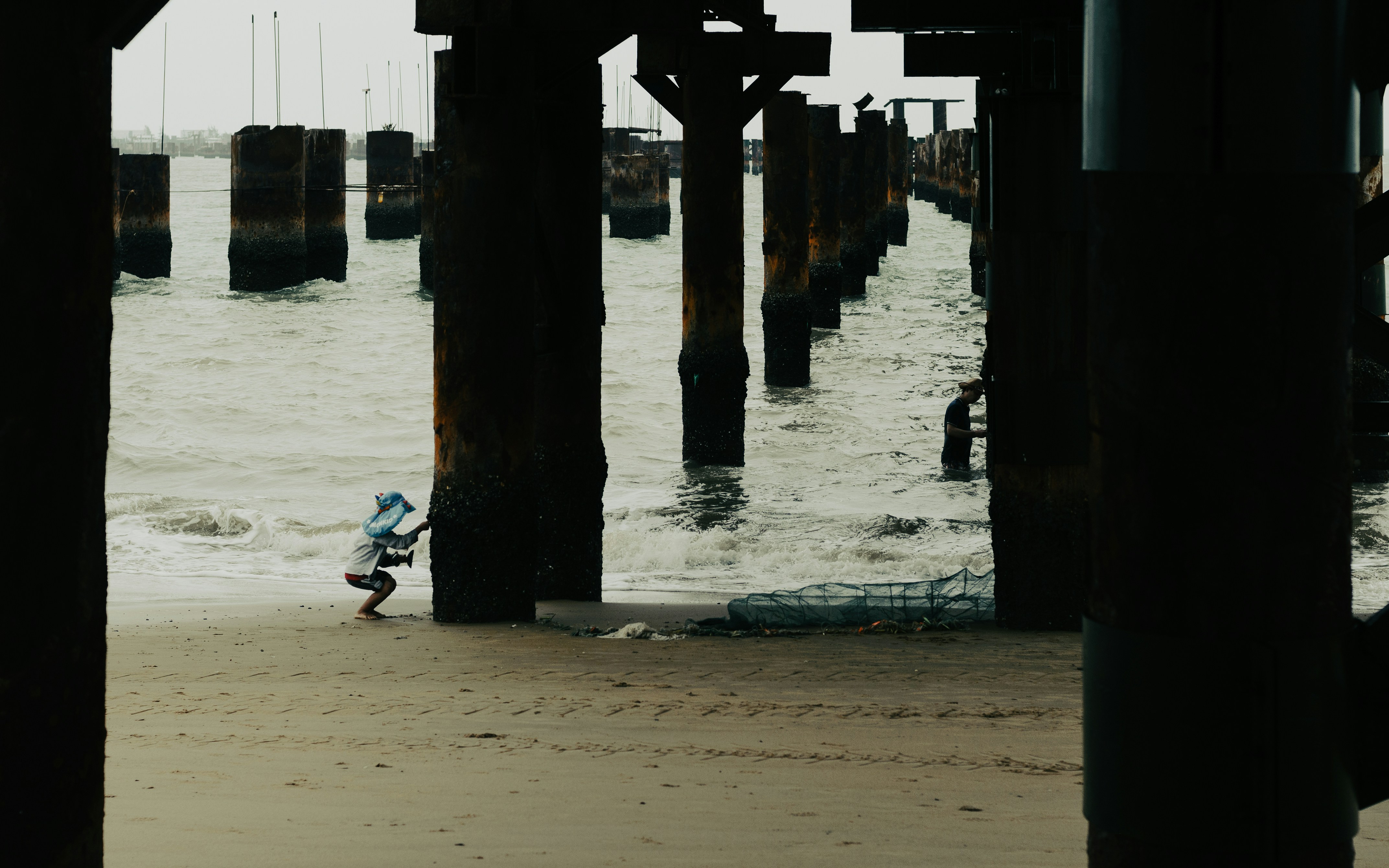 Under the Pier: A Child’s Quiet Dash Along Wet Sand Framed by Timber Columns
