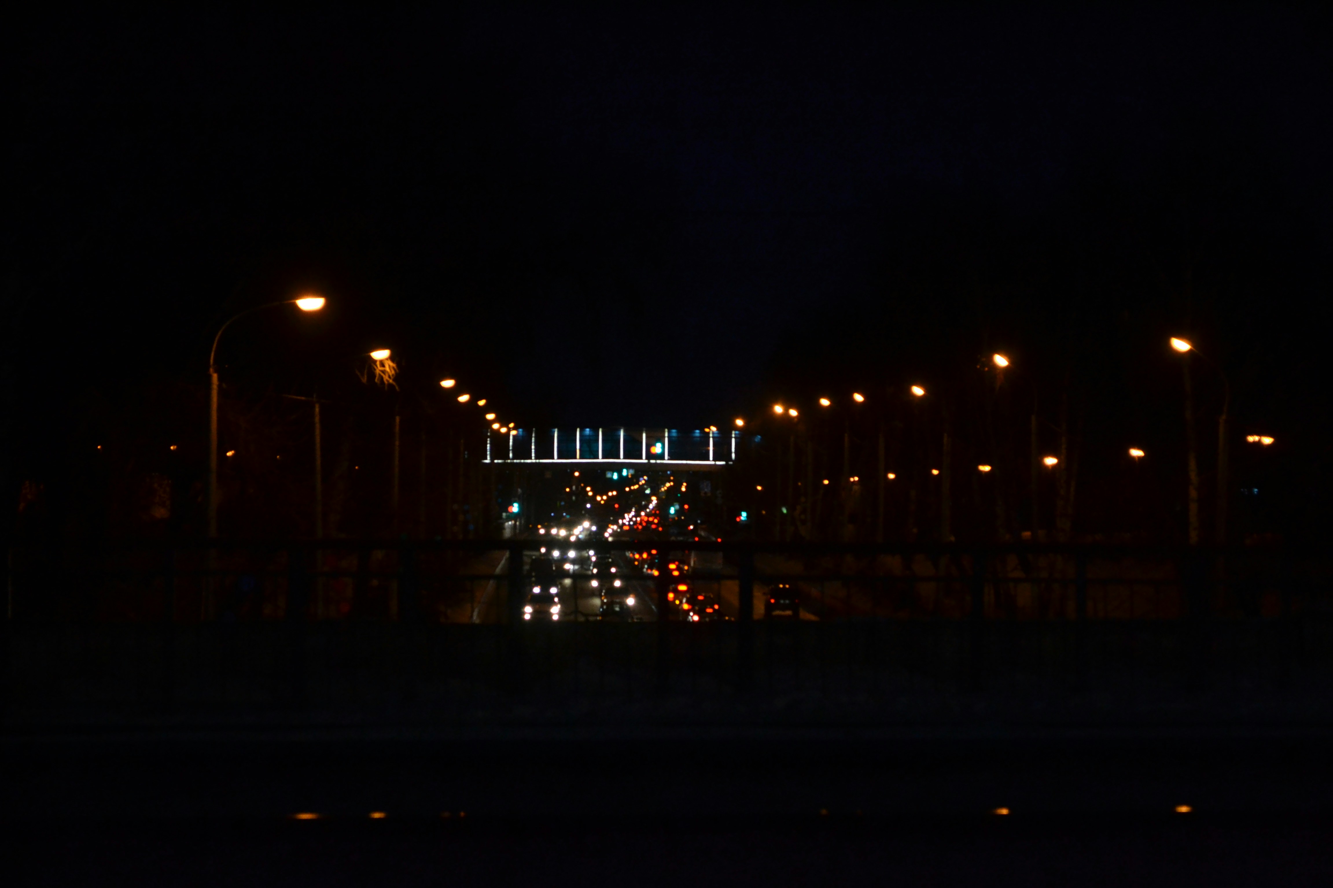 Illuminated road under a bridge at night, showcasing streaks of car headlights and streetlights reflecting on the wet pavement.