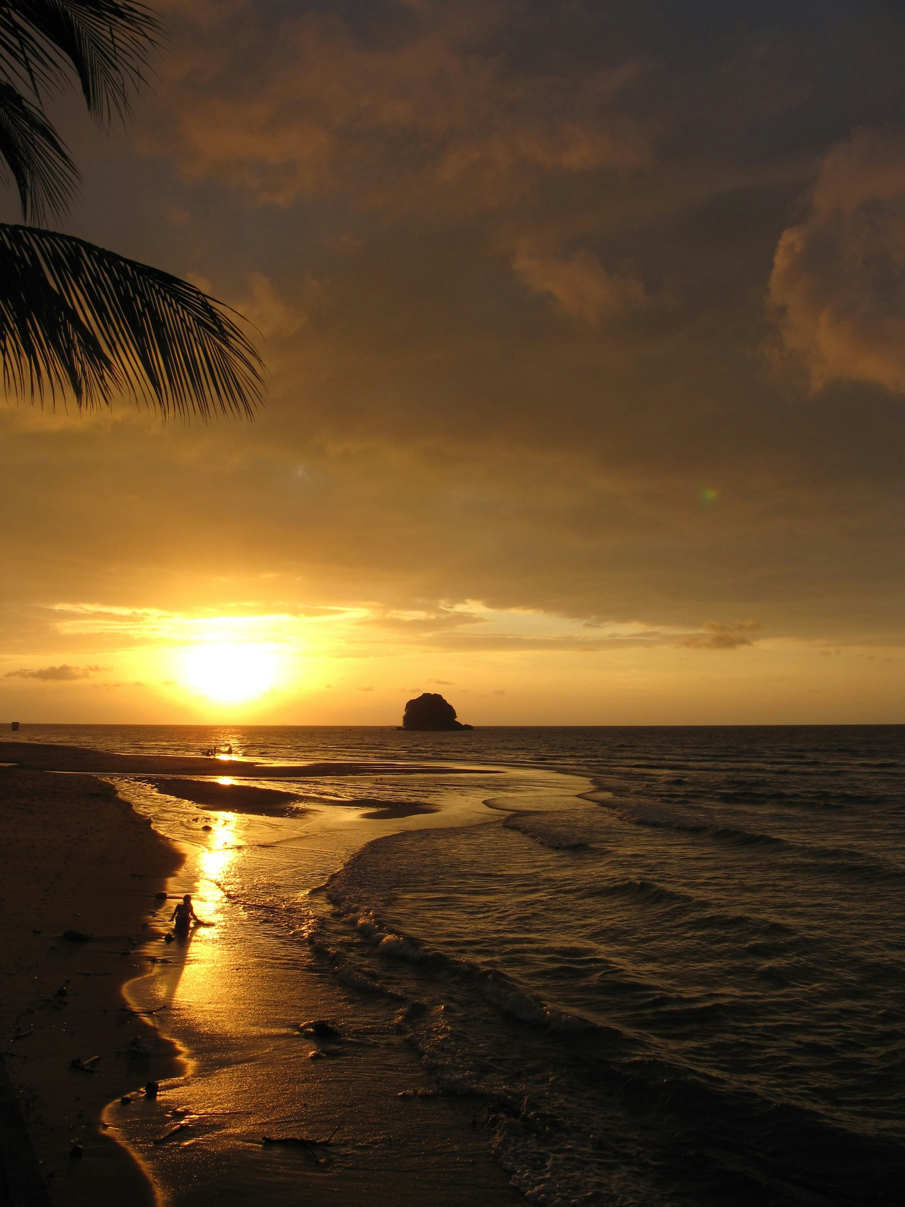 Sunset over a tranquil beach with golden reflections on the waves and a distant rock formation.