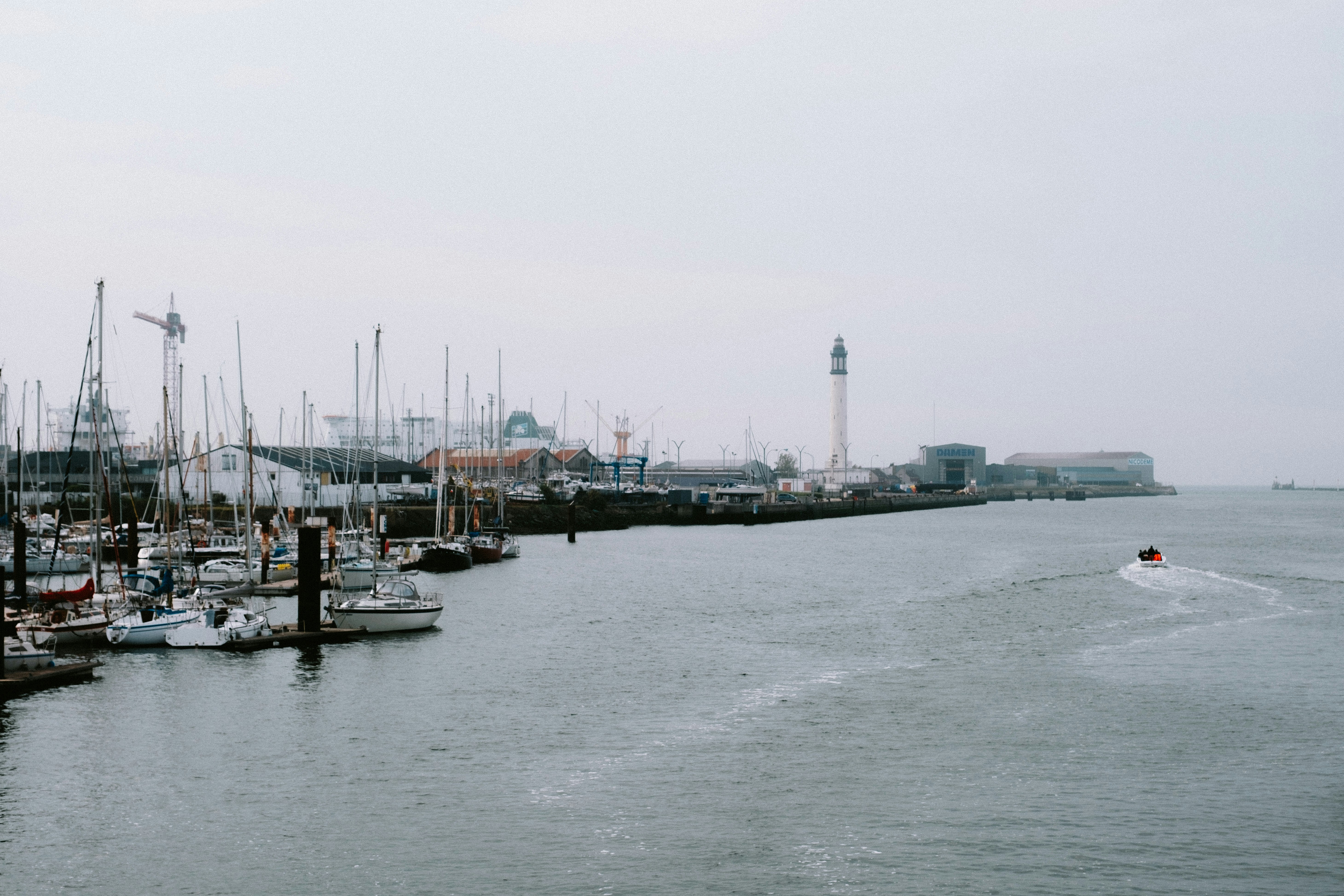 Calm harbor scene with boats docked alongside a lighthouse in the distance, under a soft gray sky.