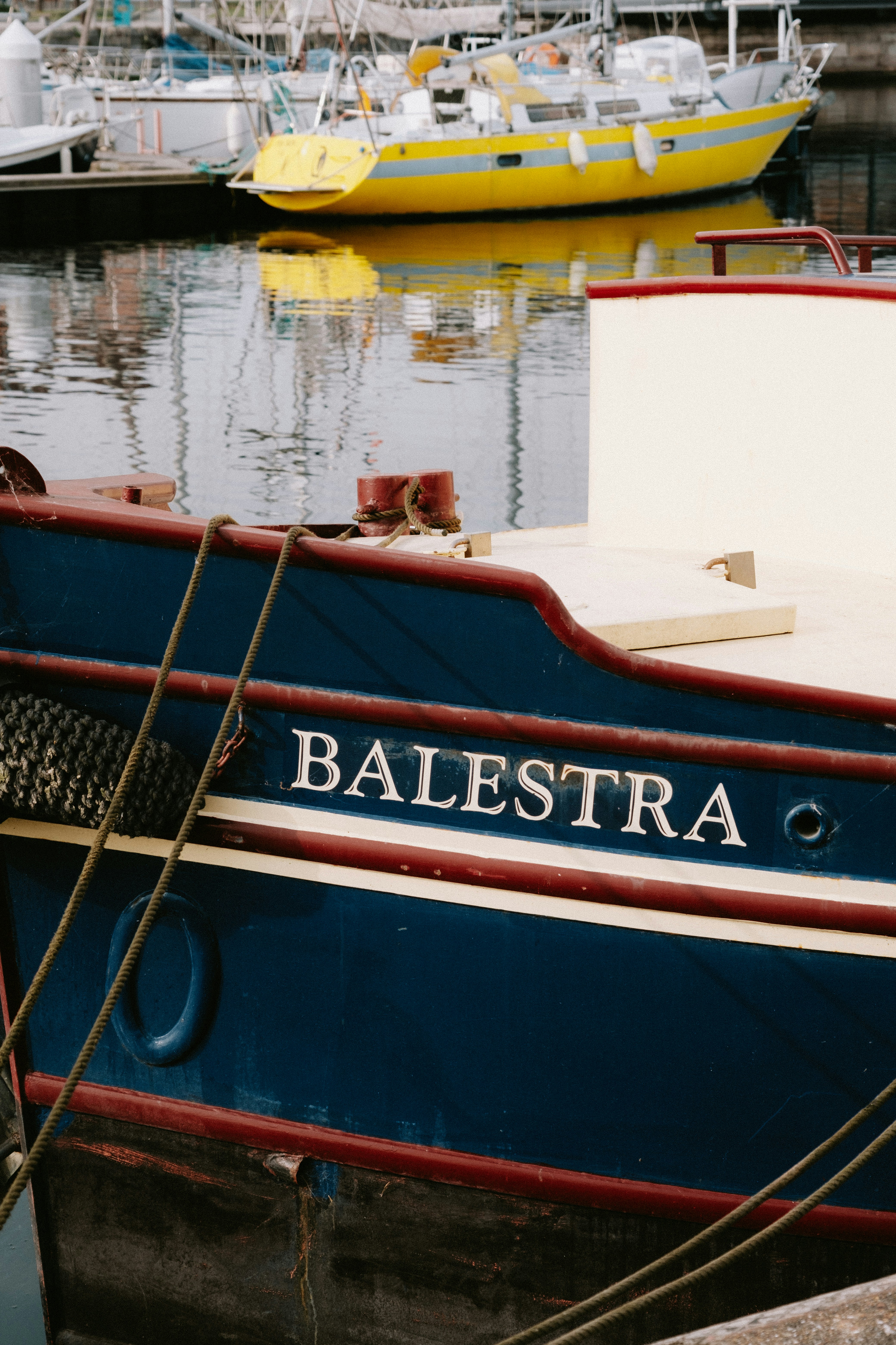 The bow of the Balestra boat adorned with its name, moored beside a vibrant yellow vessel in a tranquil harbor setting.