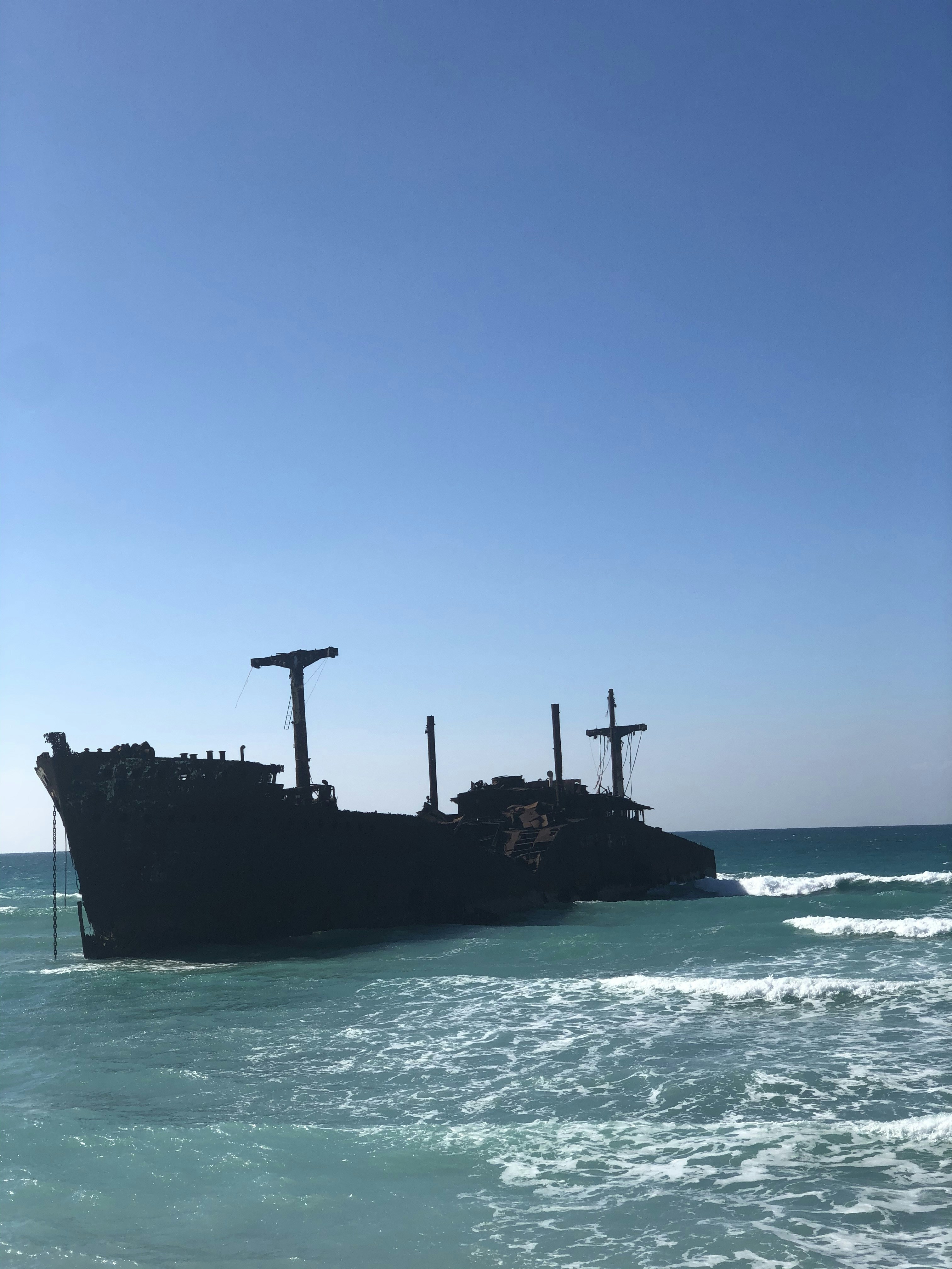 Rusting shipwreck emerging from turquoise waves under a clear blue sky. The scene captures the haunting beauty of maritime history.
