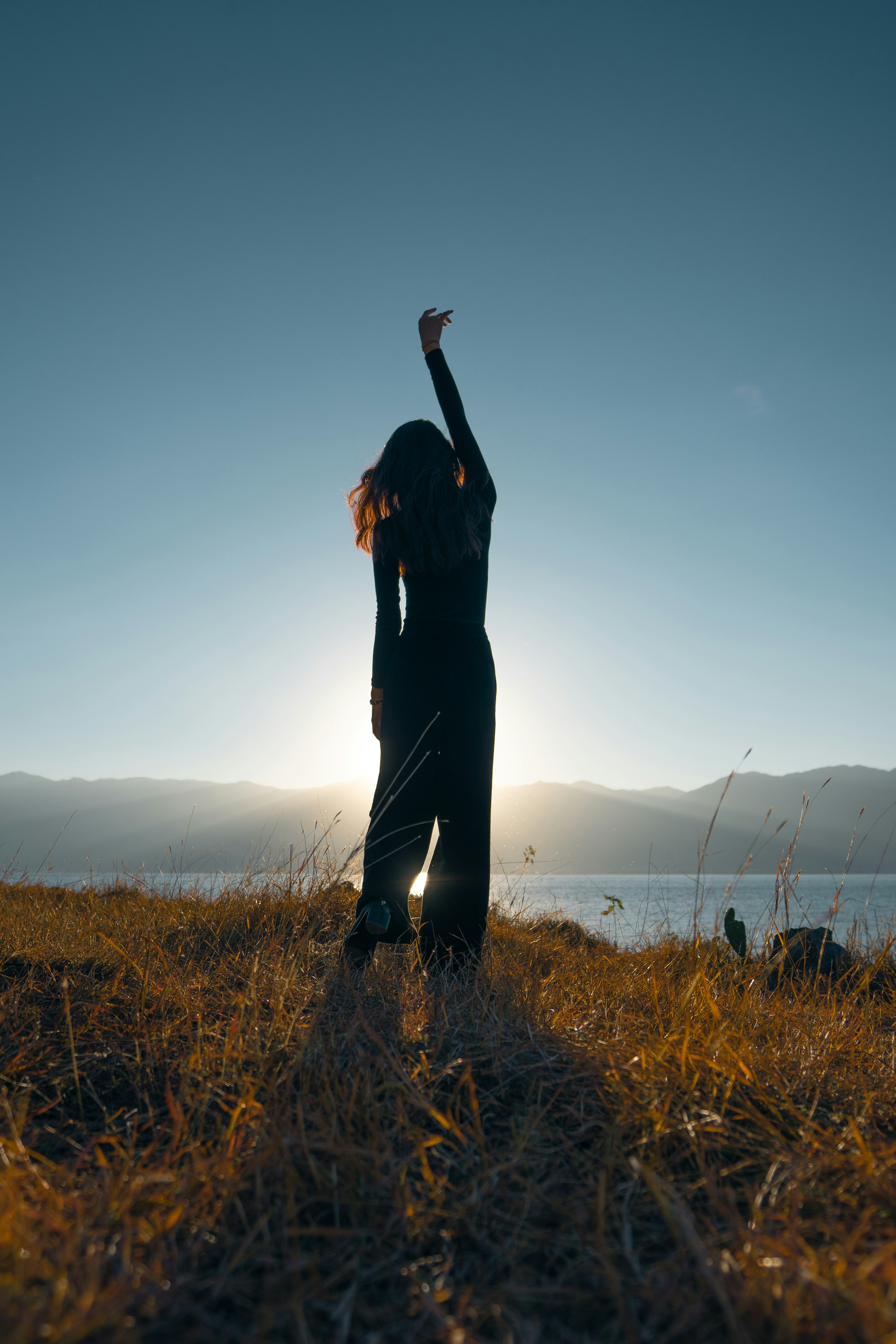 Silhouette of a person standing with one arm raised in a sunlit field, with mountains and water in the background.