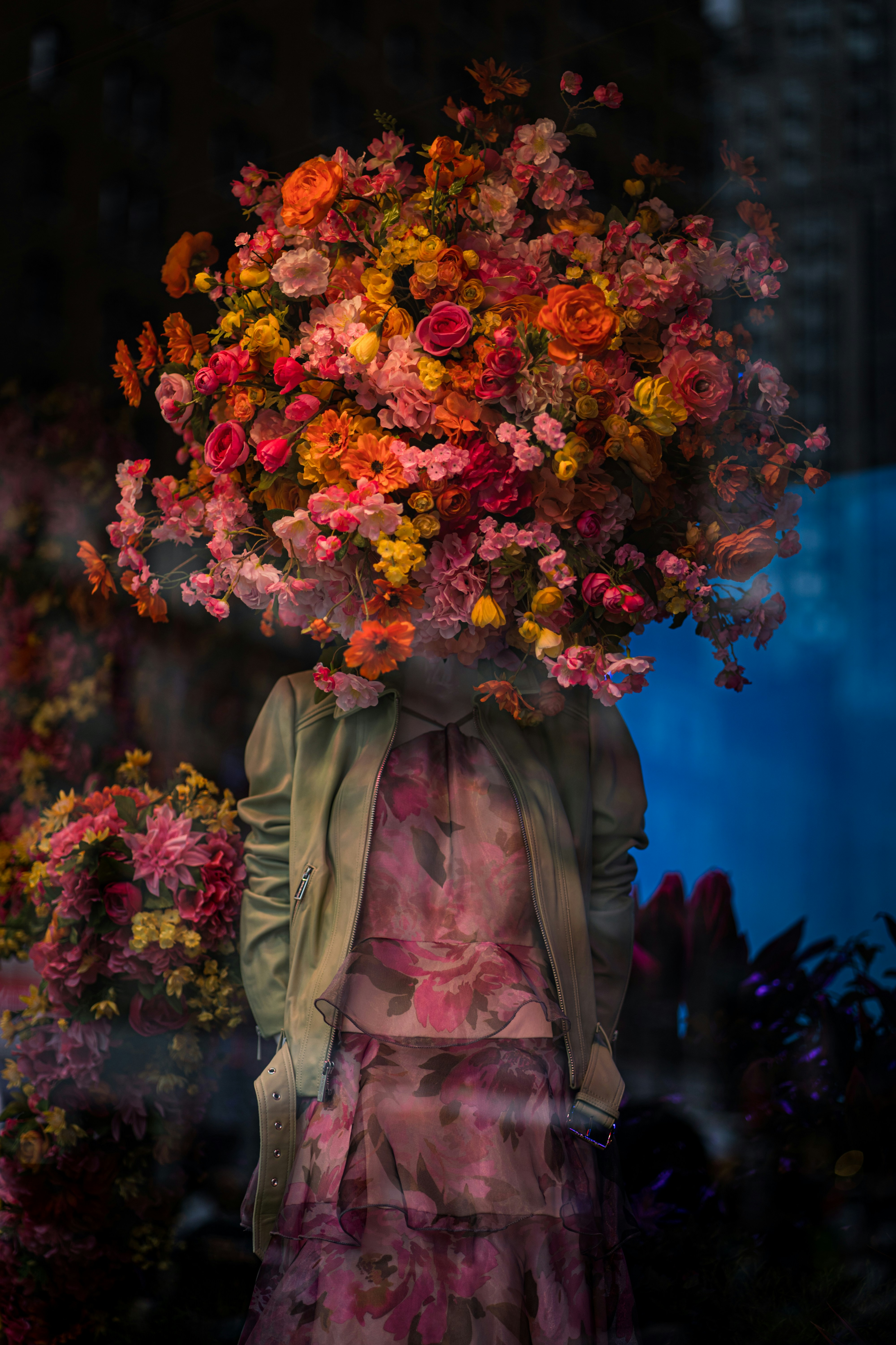 A figure adorned with a vibrant floral arrangement, obscuring their head, stands amidst a colorful backdrop of flowers. The scene blends fashion and nature, creating a surreal visual narrative.
