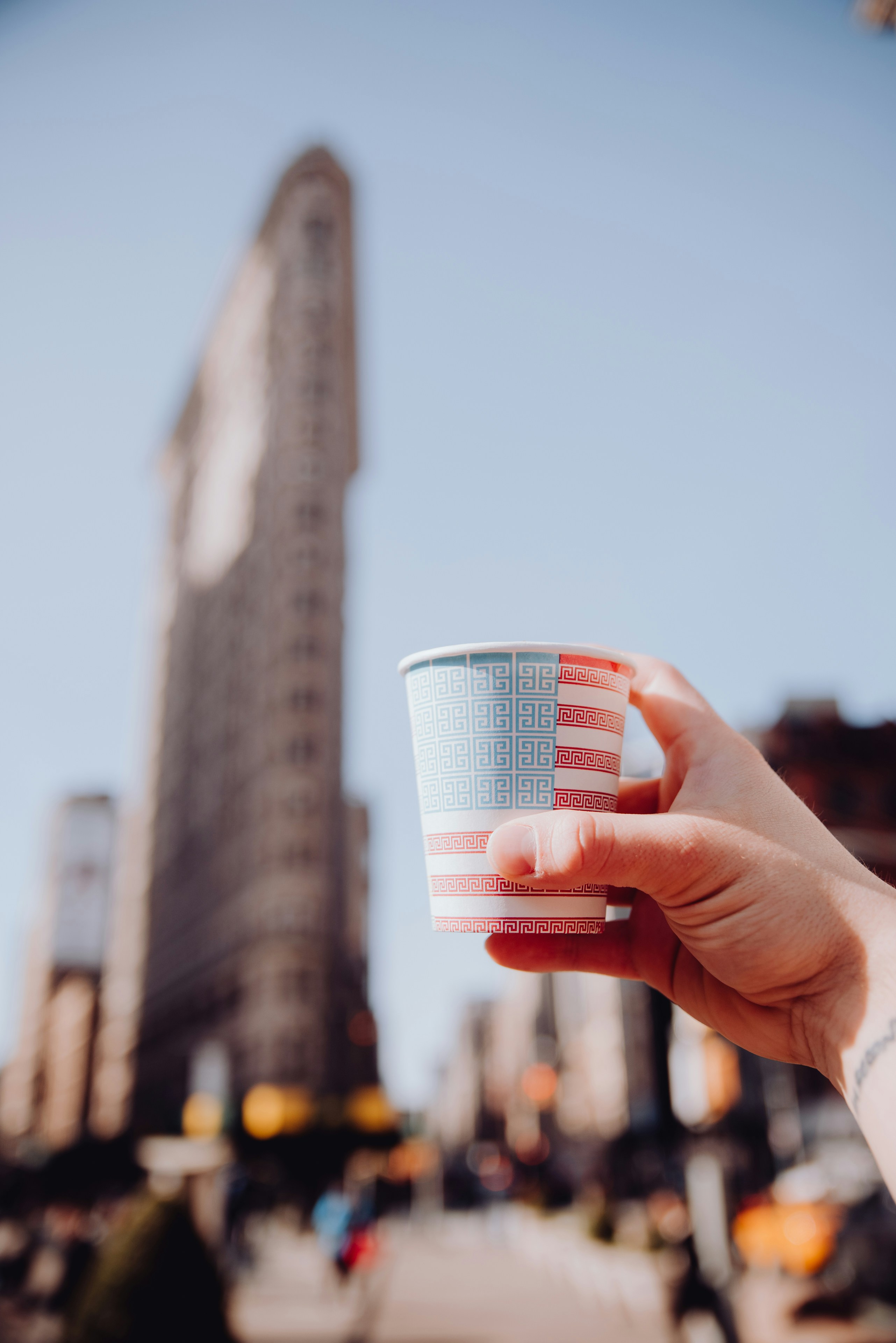 A hand holding a colorful paper cup against the backdrop of a towering building in a bustling city. The scene captures the essence of urban life.