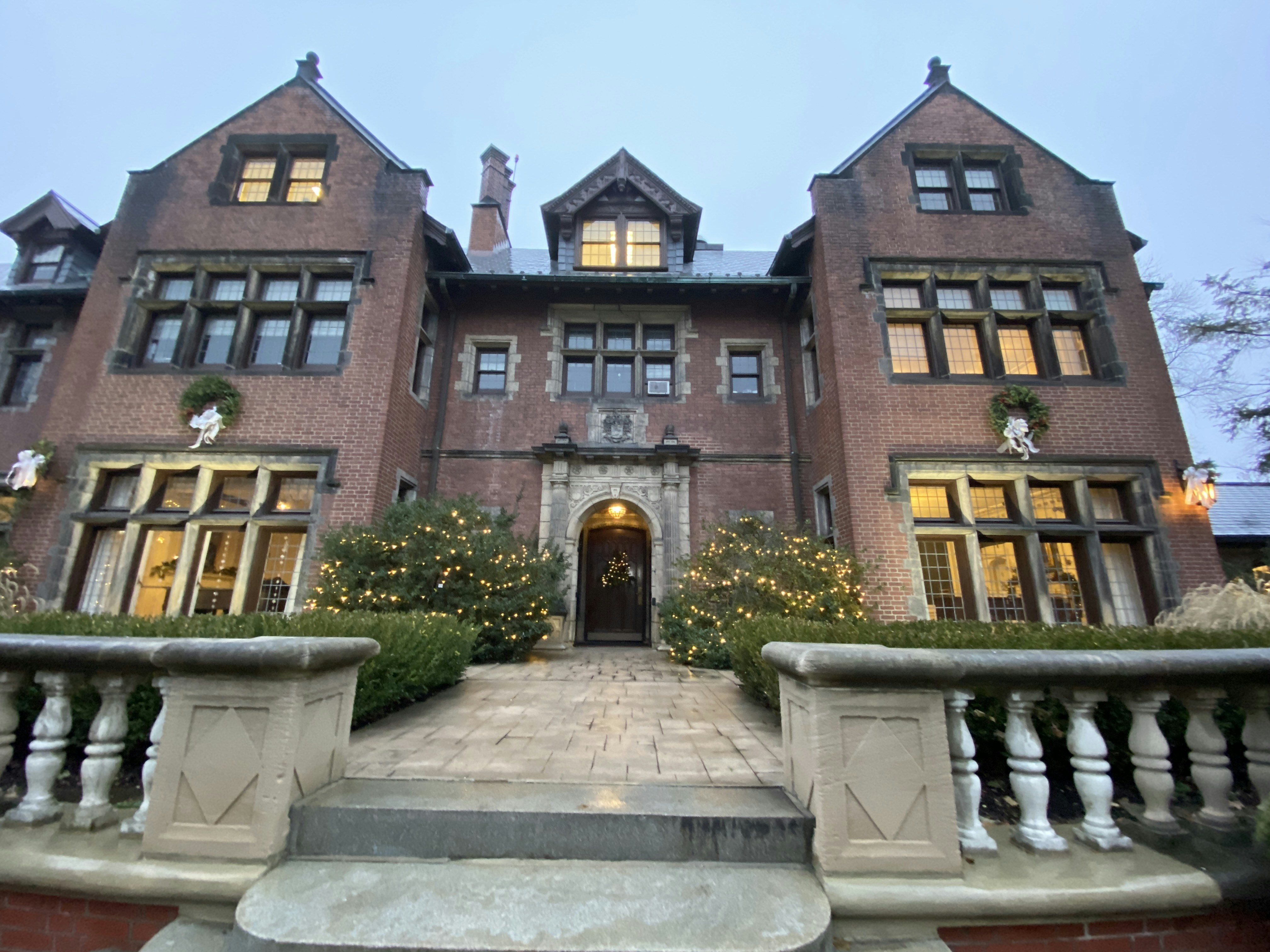 Grand brick manor adorned with holiday lights, showcasing intricate architecture and lush greenery in the foreground.