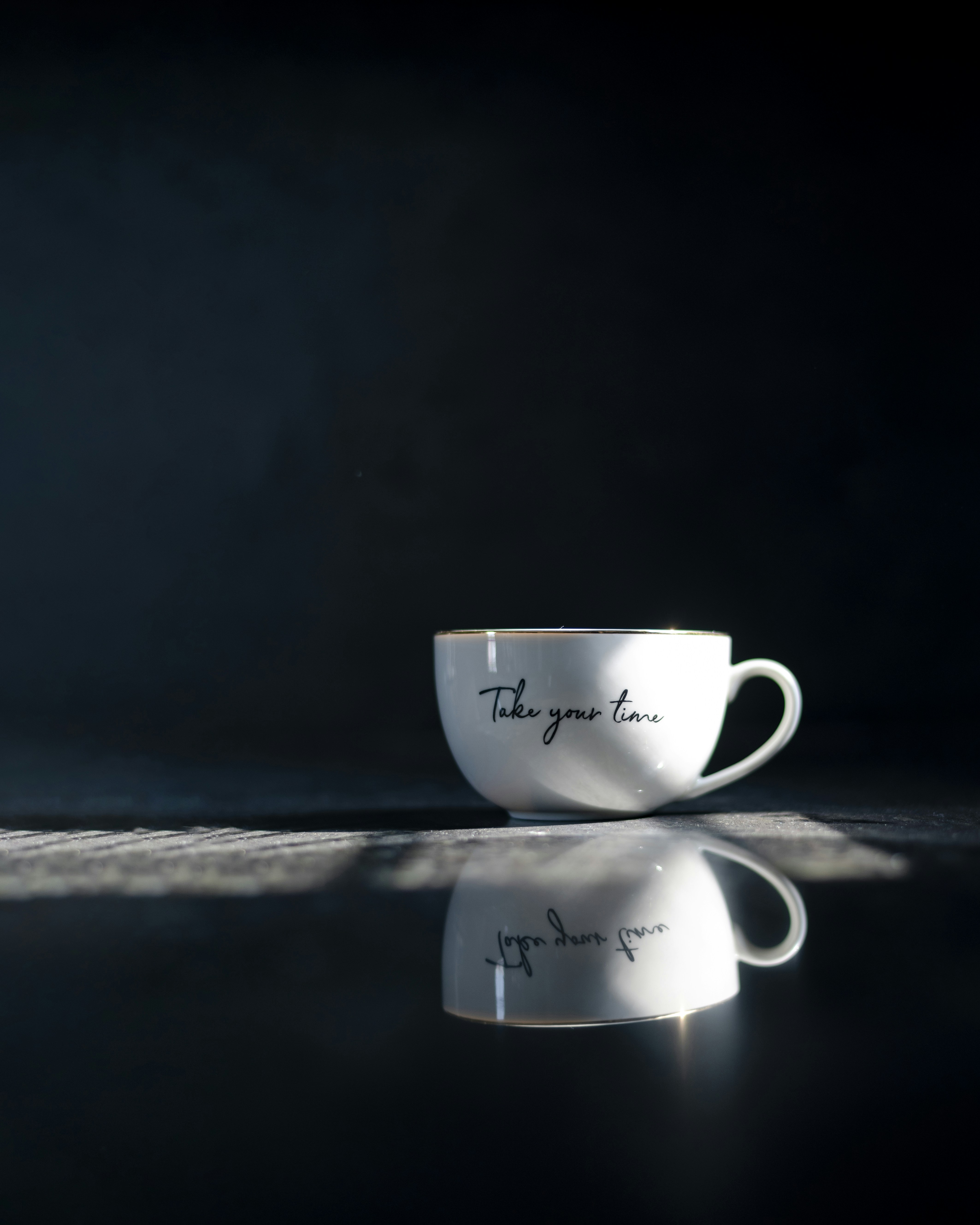 White ceramic cup with the phrase 'Take your time' resting on a reflective surface, illuminated by soft light against a dark backdrop.