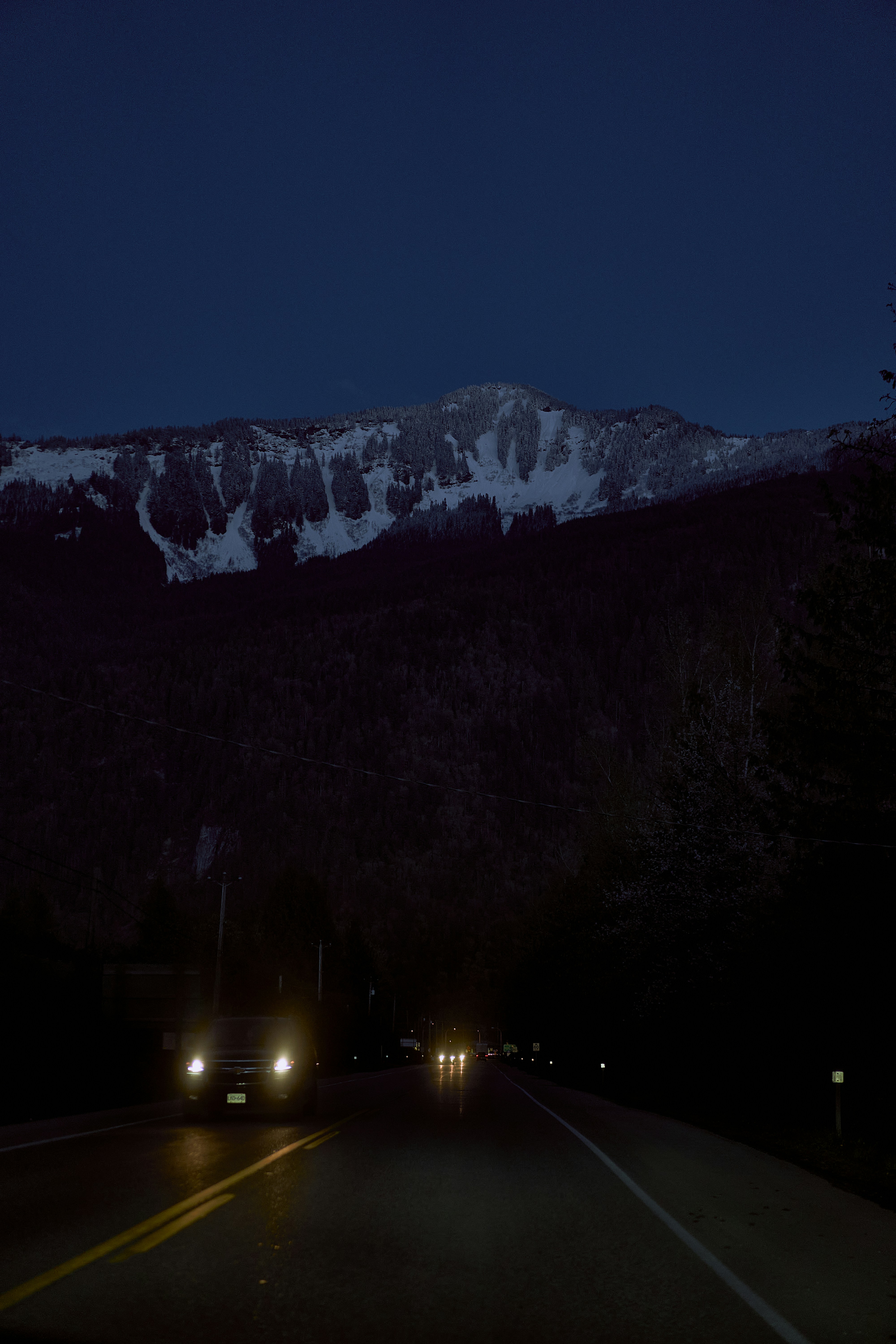 A dimly lit mountain road under twilight, with headlights illuminating the path and a snow-capped peak looming in the background.