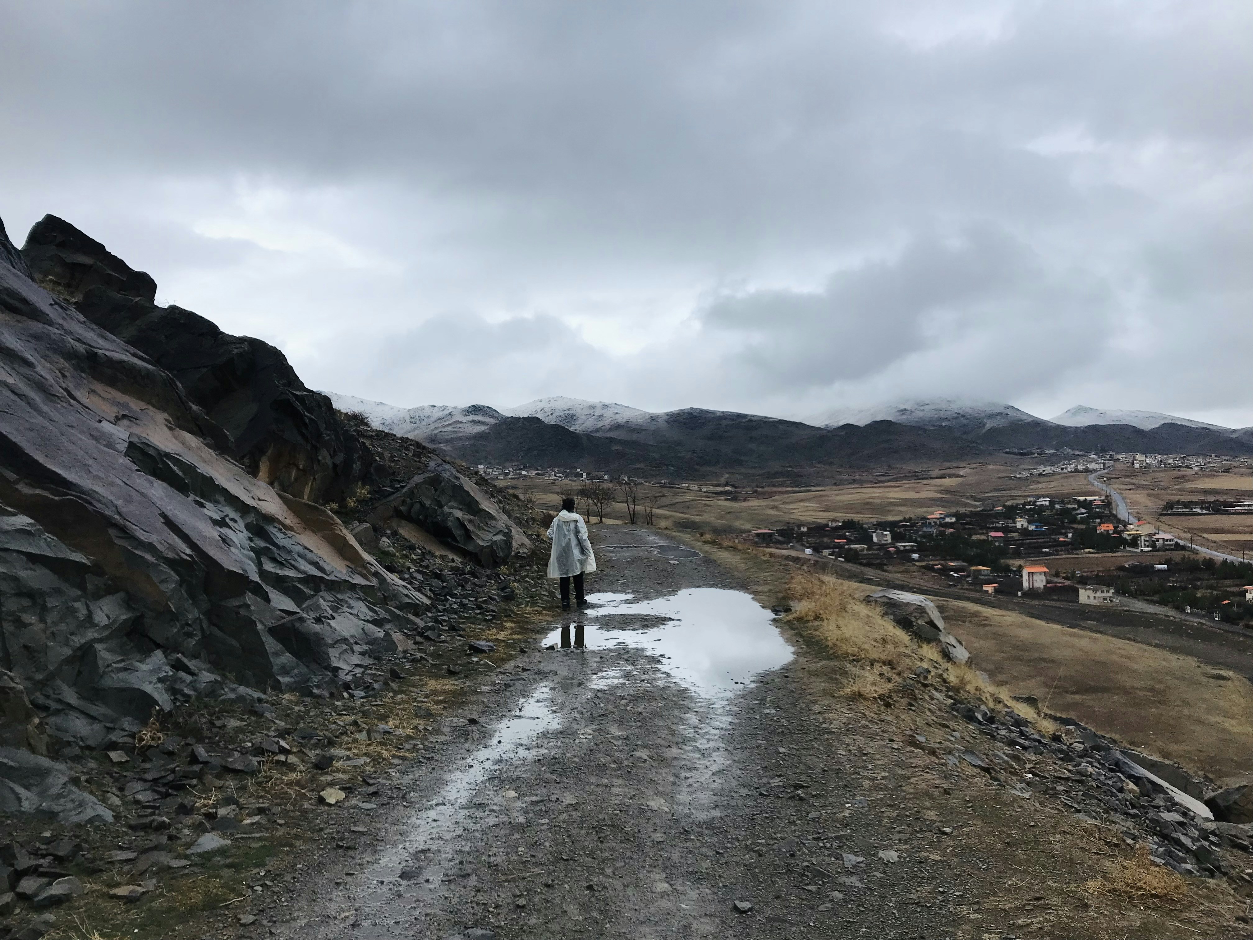 Person walking along a rocky dirt road under overcast skies with distant mountains in view.