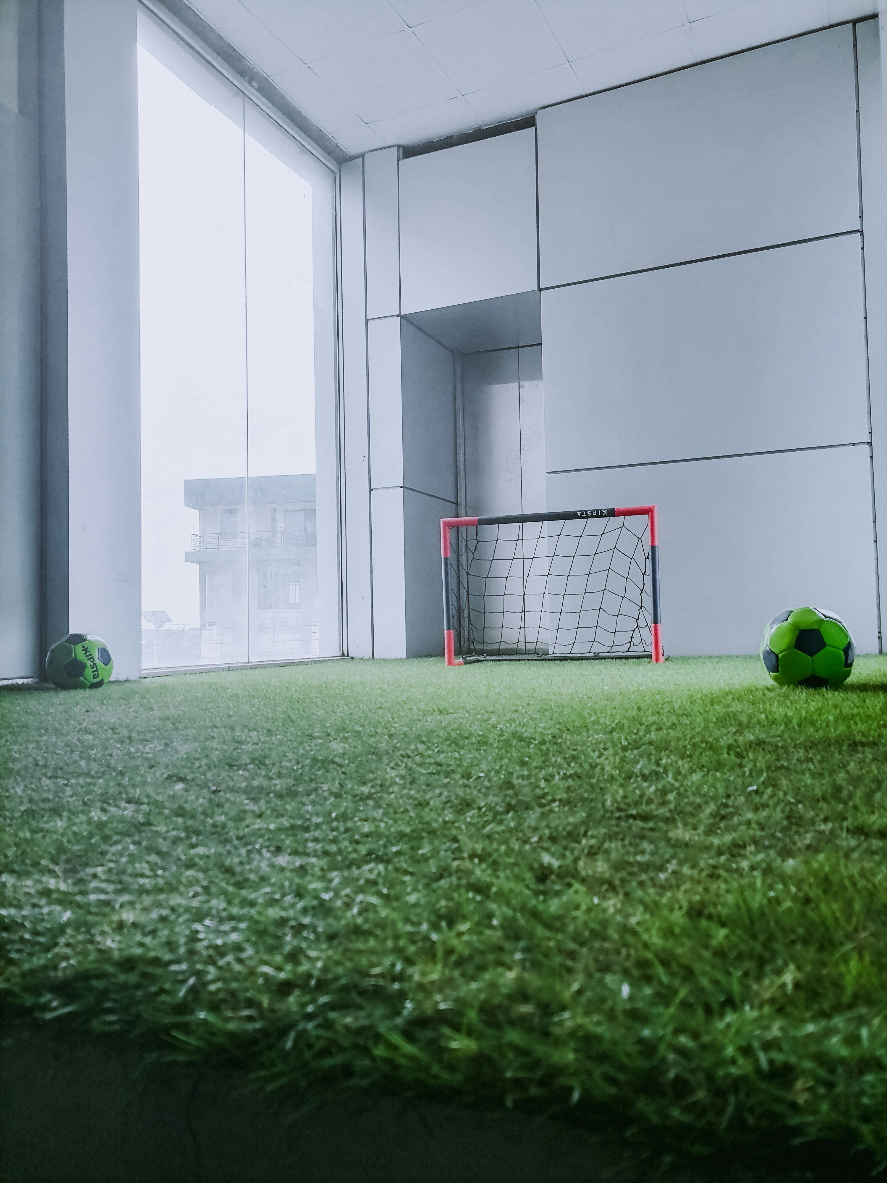 Indoor soccer field featuring a small goal and two soccer balls on artificial grass, framed by large windows allowing natural light to filter in.