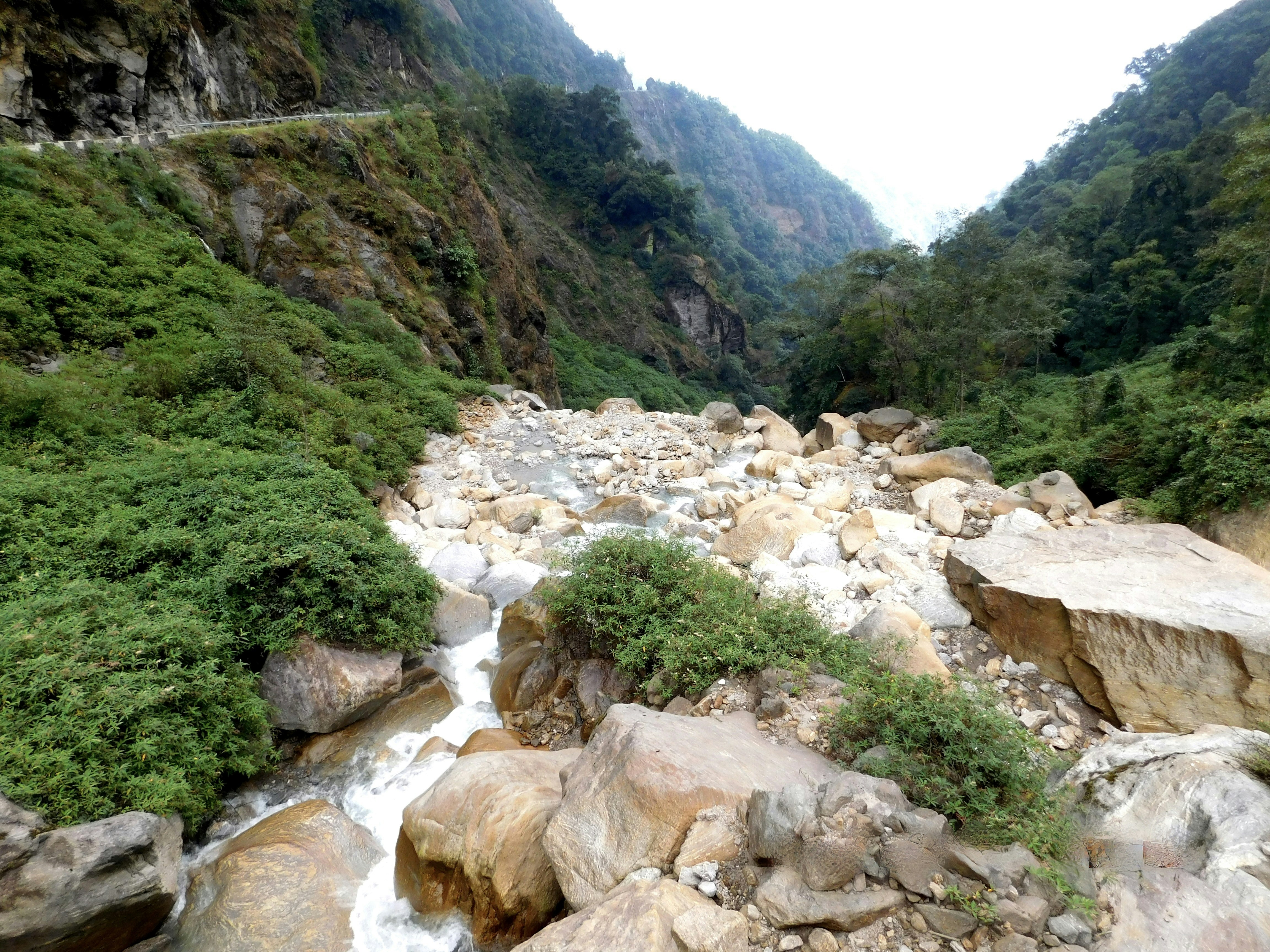 A rocky riverbed flows through a lush green valley, framed by steep mountains under a hazy sky.