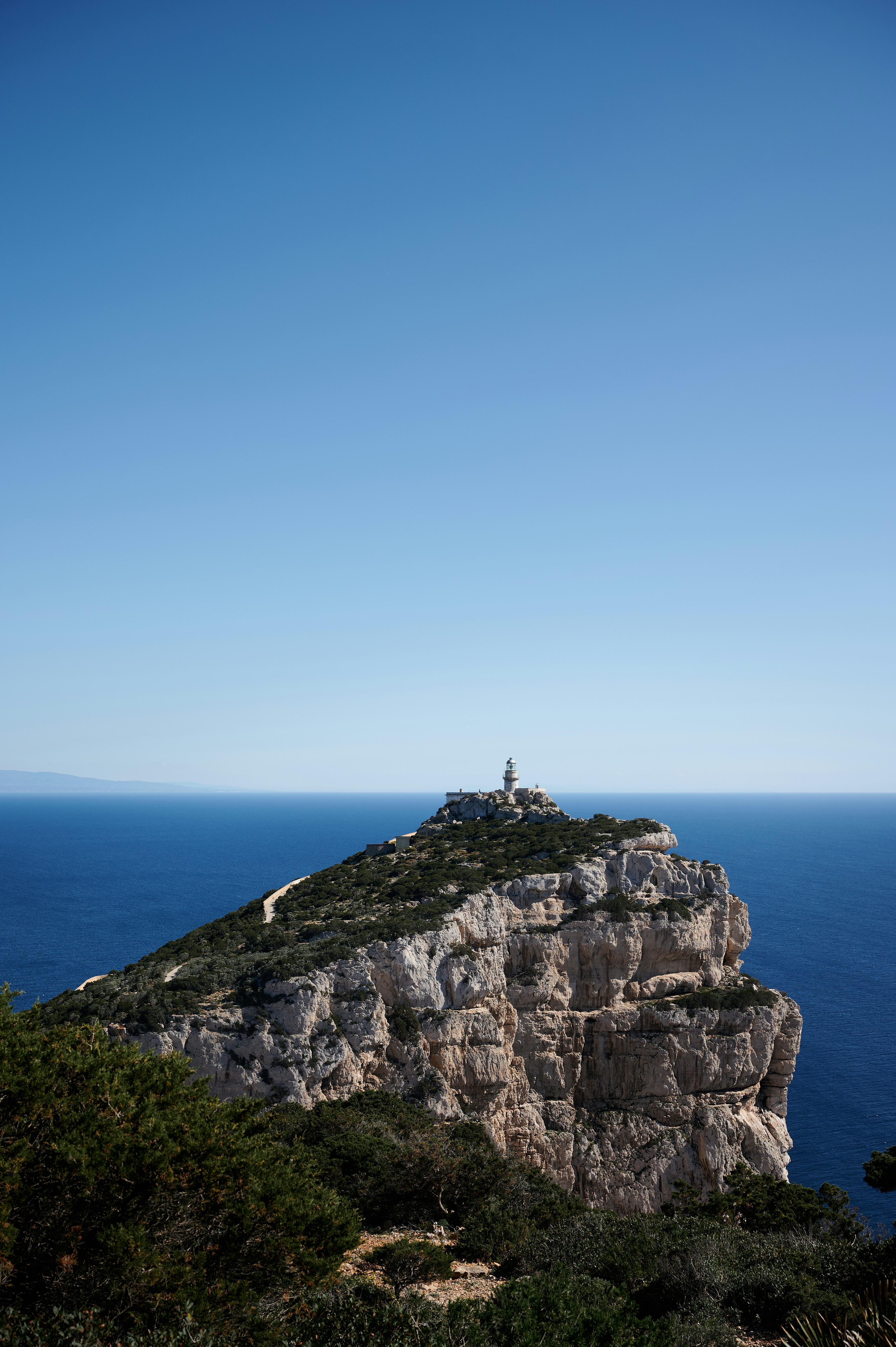 A historic lighthouse perched atop a rugged cliff, overlooking a vast expanse of deep blue ocean under a clear sky.