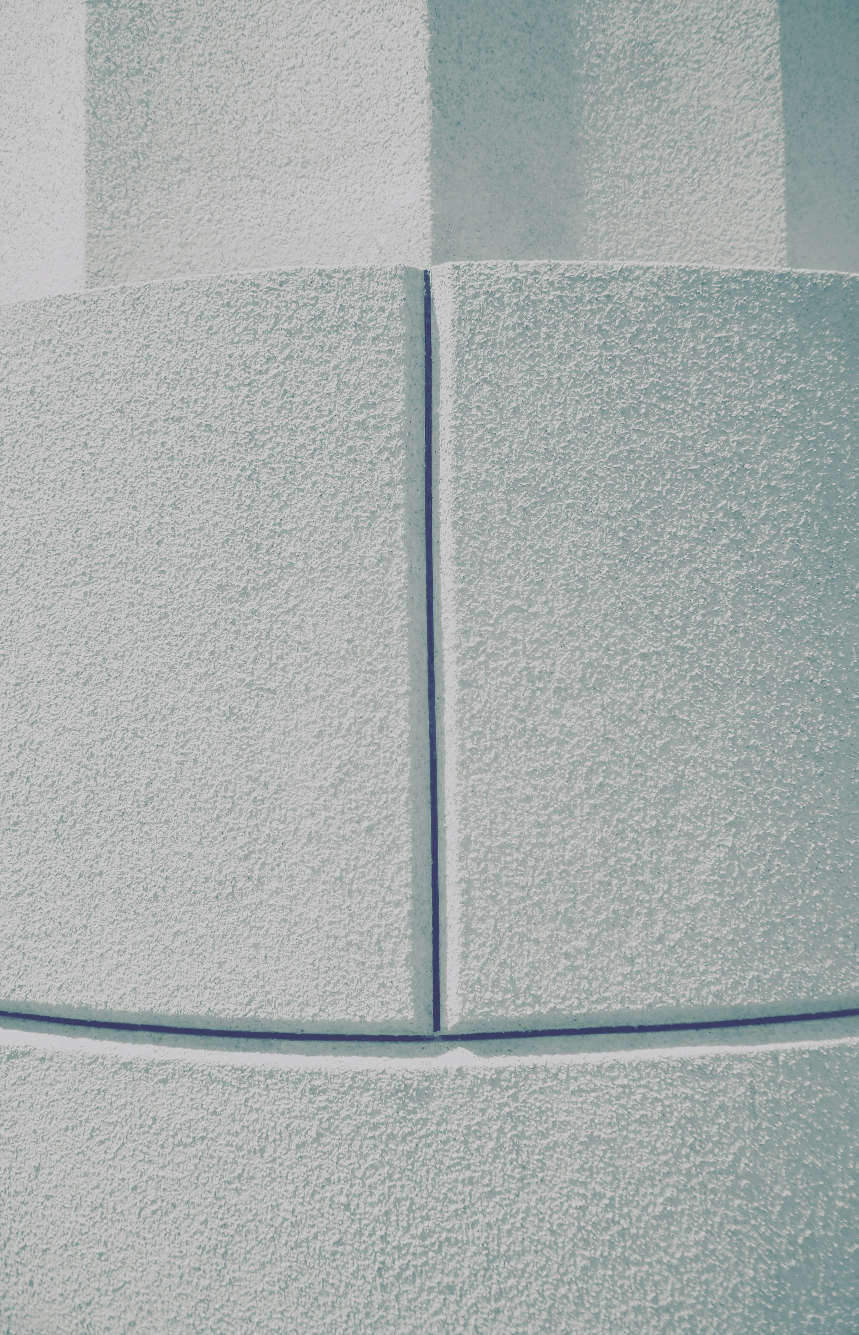 Close-up of pale textured concrete panels with fine stippling and curved seam lines, creating an abstract architectural pattern. The image emphasizes material texture and geometry without a dominant subject.