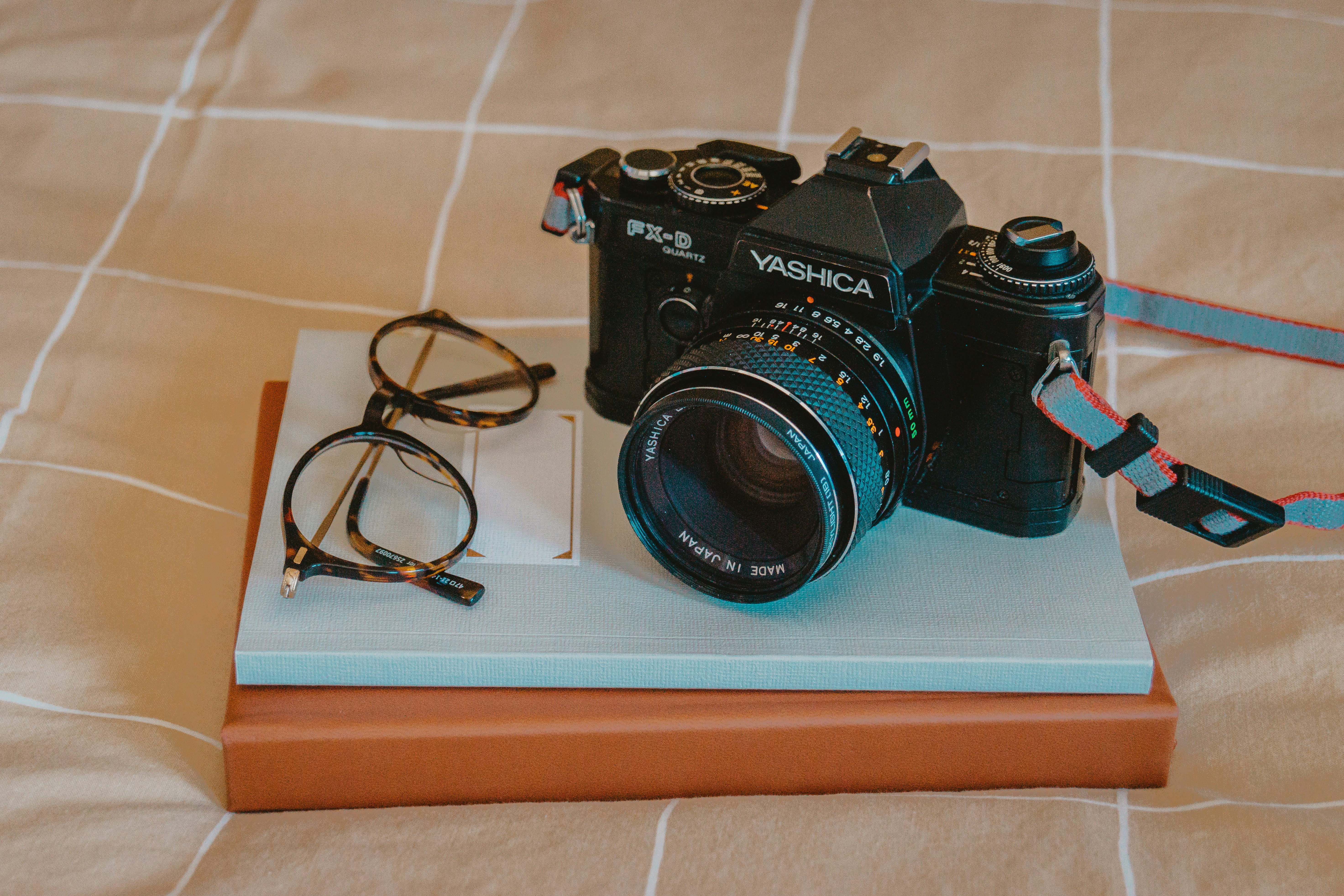 A vintage Yashica film camera rests on two stacked books with a pair of round glasses nearby on a striped bedspread.