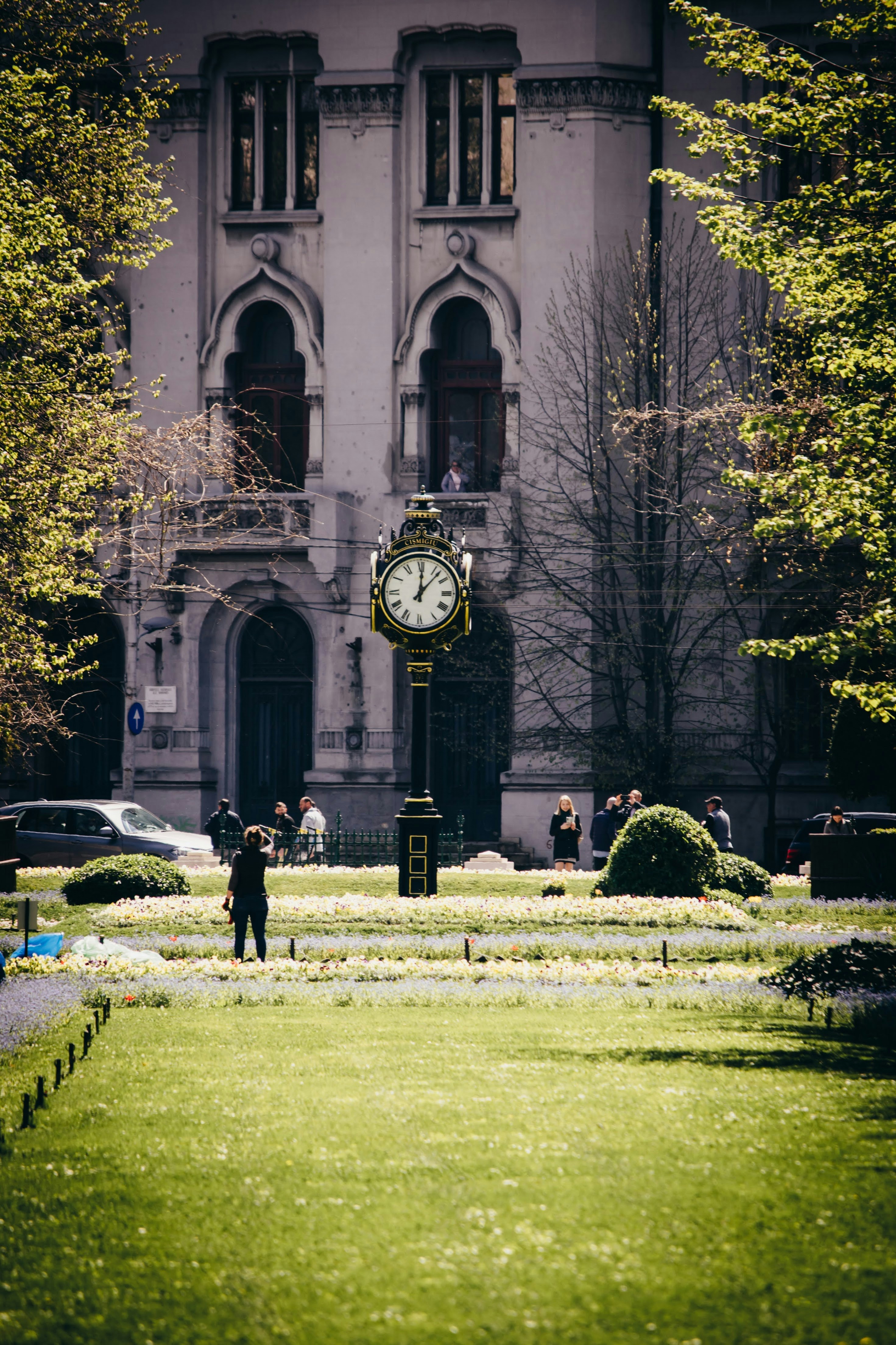 A vintage clock stands prominently in a lush garden, surrounded by blooming flowers and visitors enjoying the scenery.