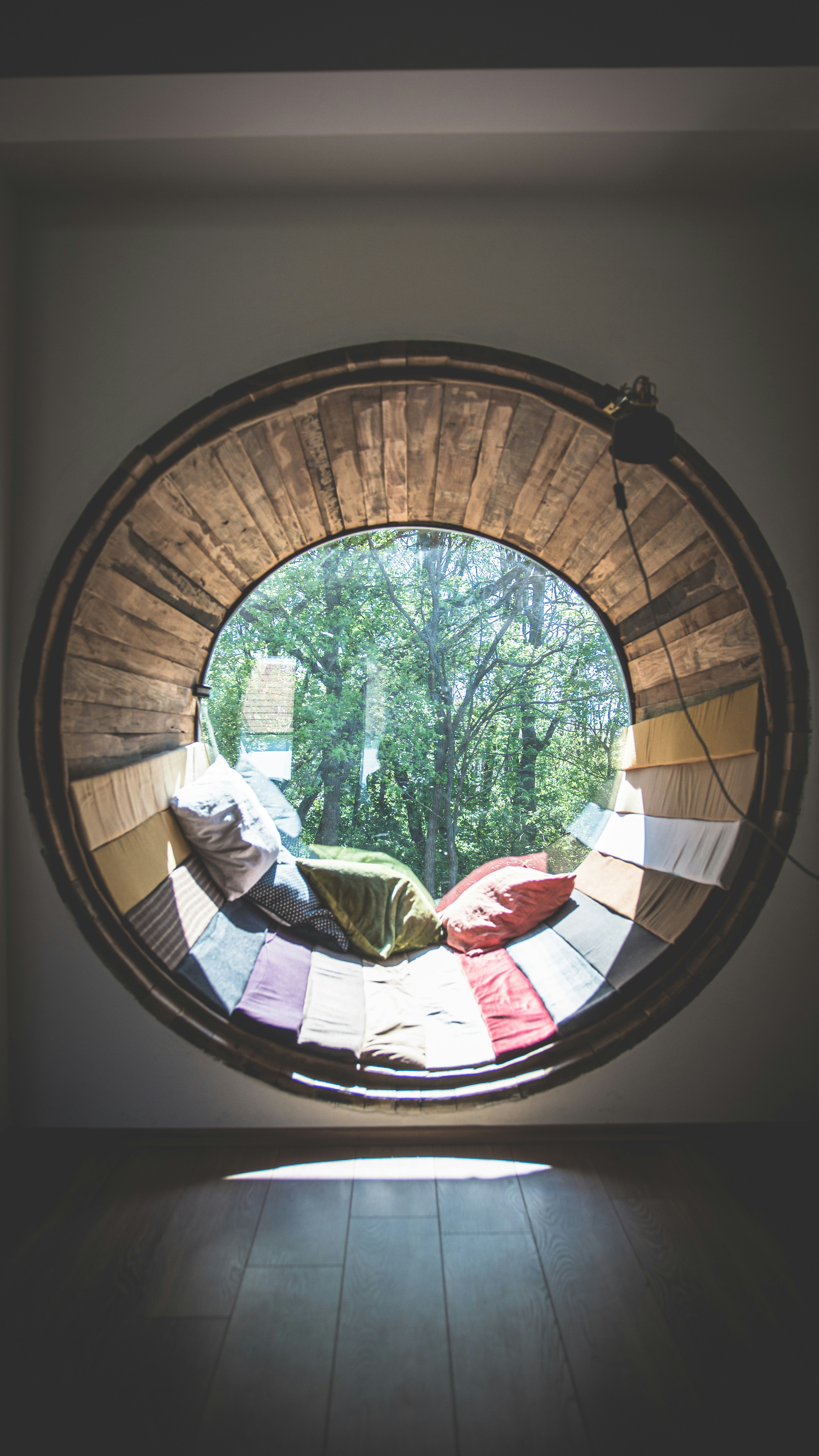 Circular wooden window frame with colorful cushions, offering a cozy seating area that overlooks a lush green landscape.