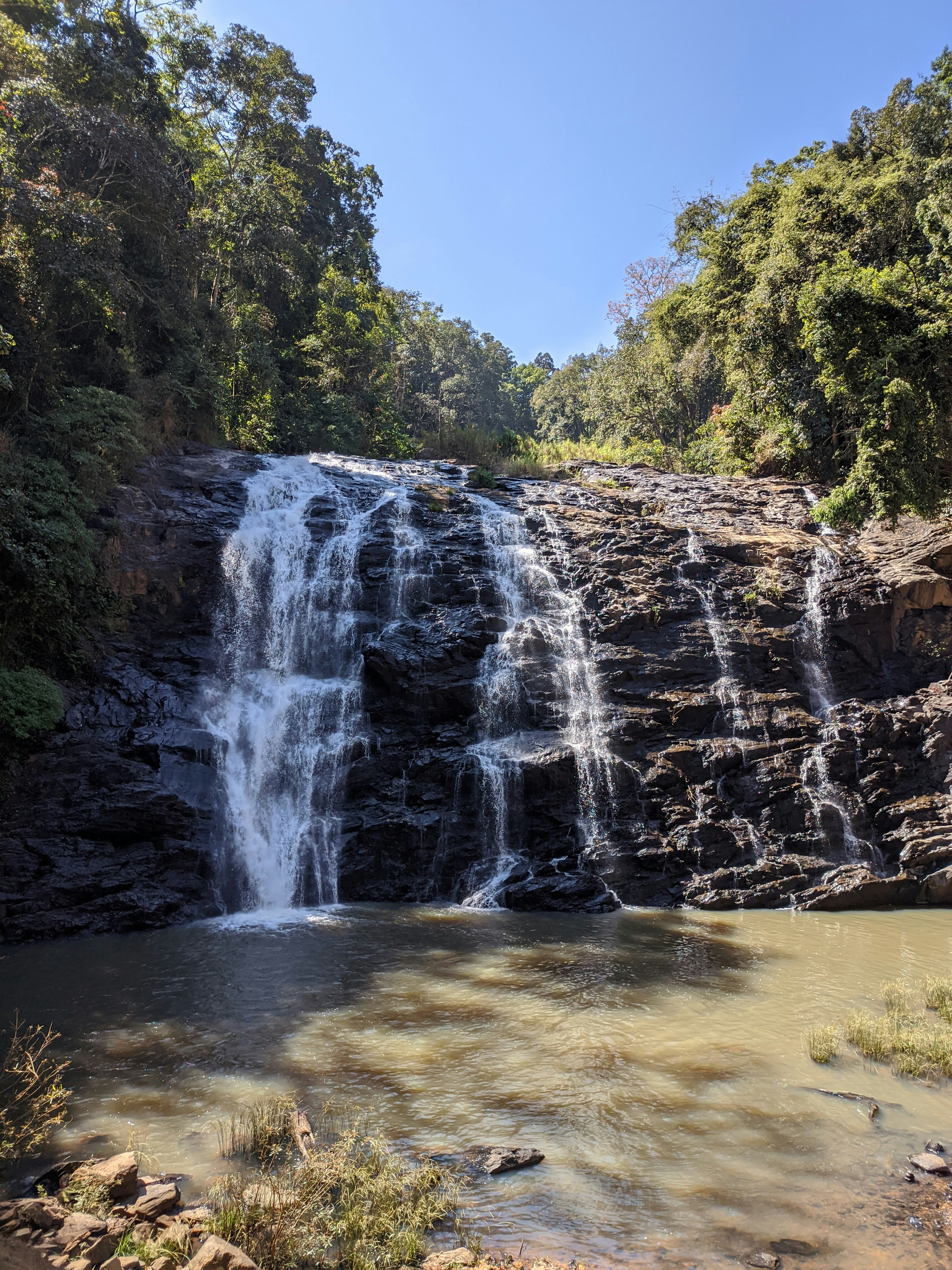 A sunlit waterfall spills over a layered rock ledge into a calm pool, framed by dense green trees under a clear blue sky.
