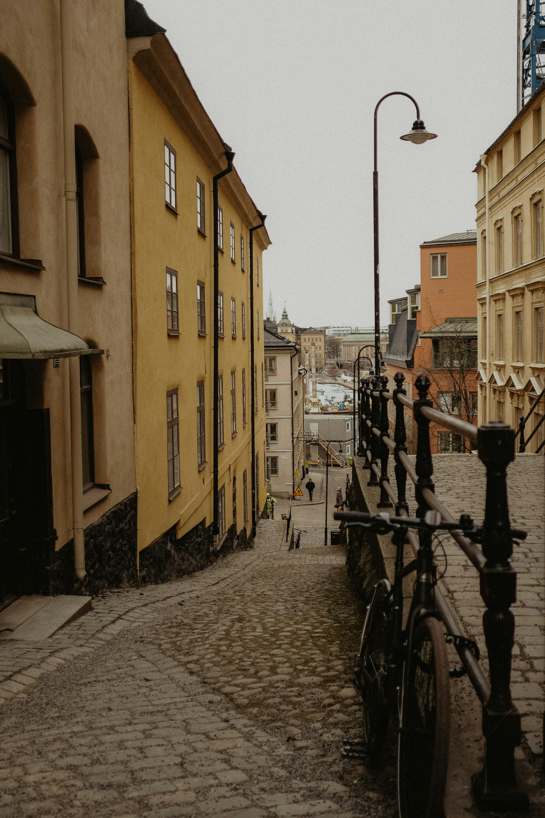 Narrow cobblestone street lined with historic buildings, leading toward a distant view of a bustling harbor. A bicycle rests against the railing, adding charm to the scene.
