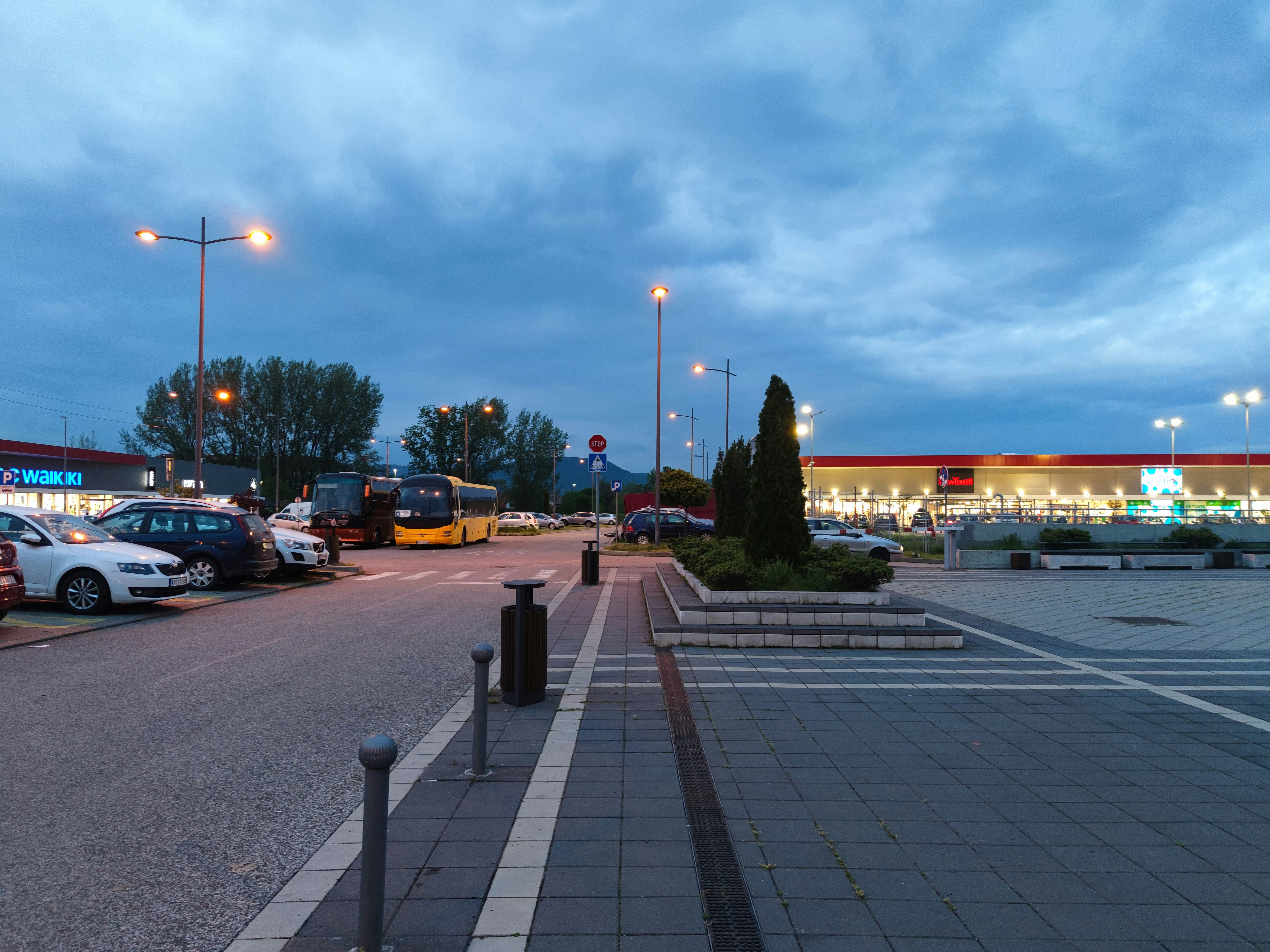Busy parking lot at twilight, with vehicles and a bus waiting near a shopping center illuminated by artificial lights.